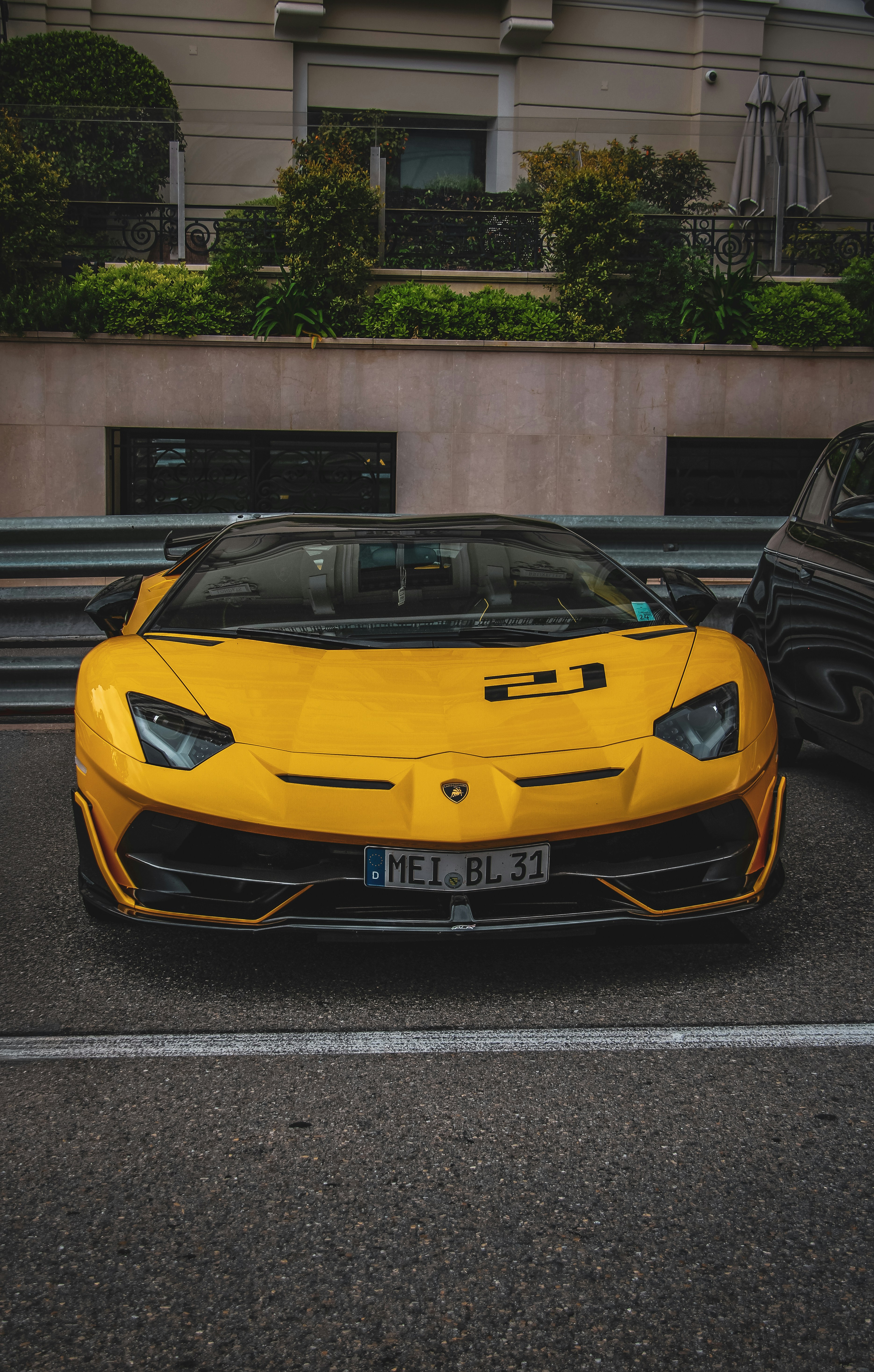 Bright yellow Lamborghini parked on a street with a building and greenery in the background. Aiz car's aggressive front and license plate are clearly visible.
