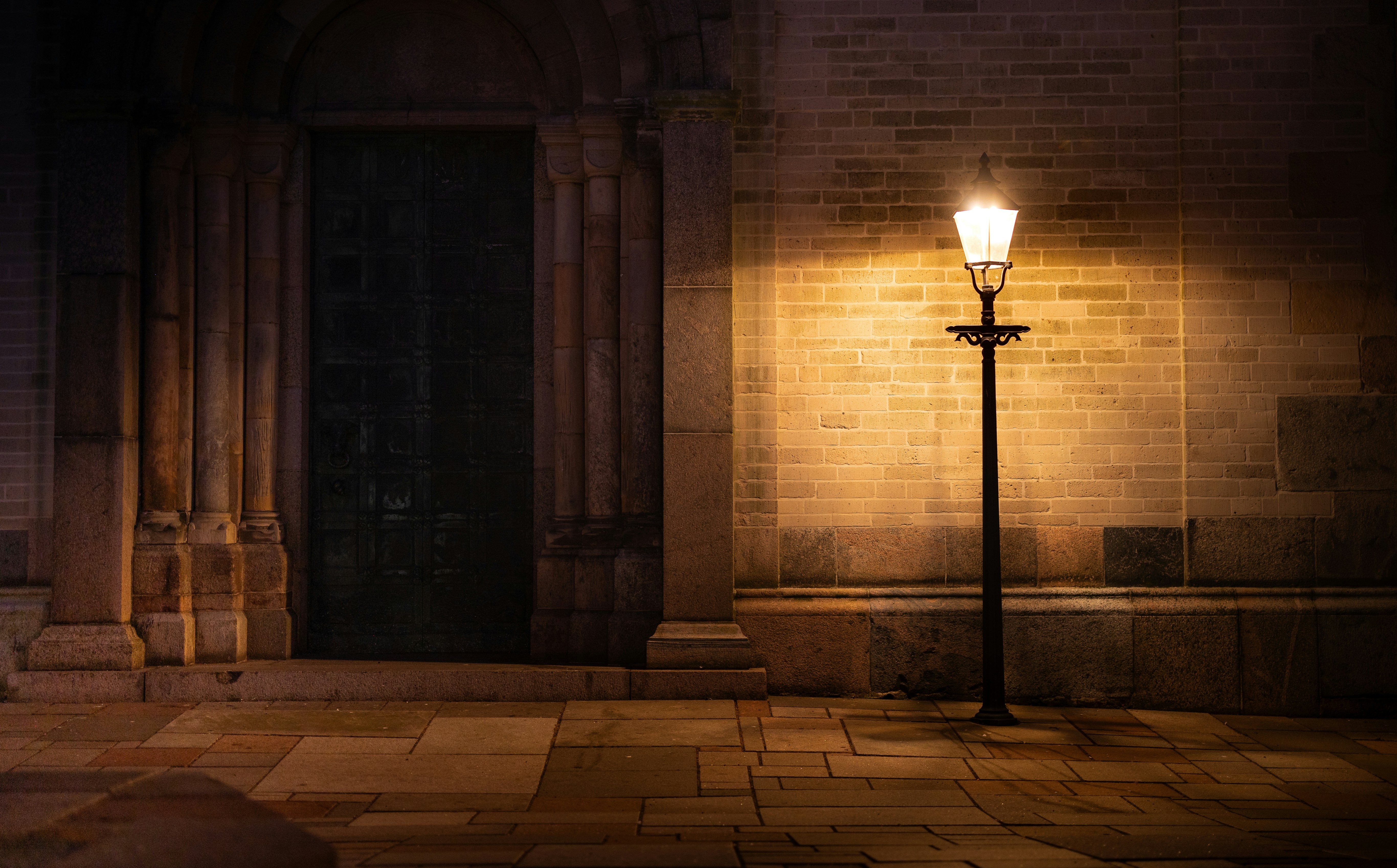 A solitary streetlamp casts a warm pool of light on a brick building and cobblestone plaza, creating a quiet nocturnal atmosphere. Note: a realistic photograph, not a painting or illustration.