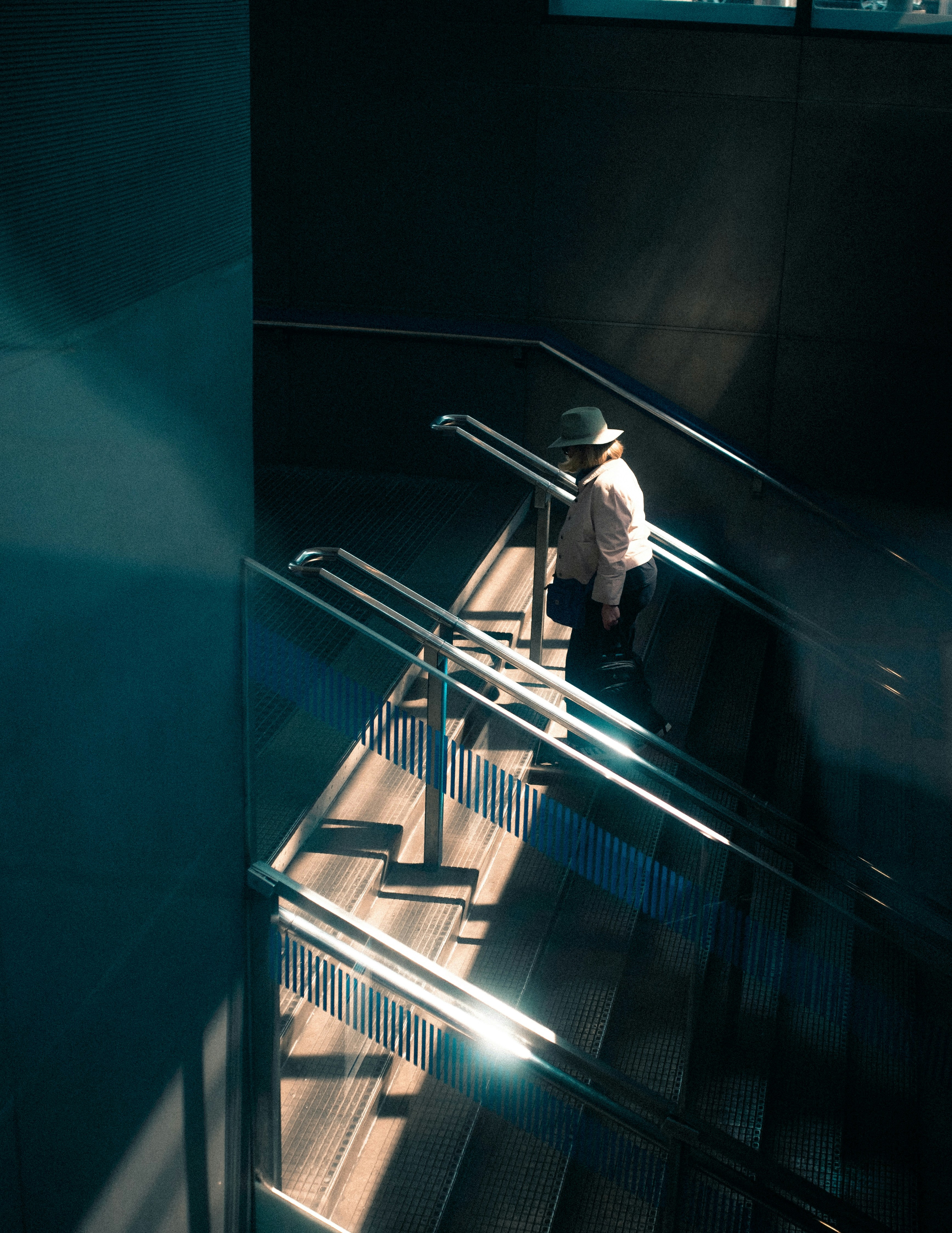 A lone traveler wearing a hat ascends a blue-toned stairwell with metallic railings, bathed in dramatic lighting and shadows.