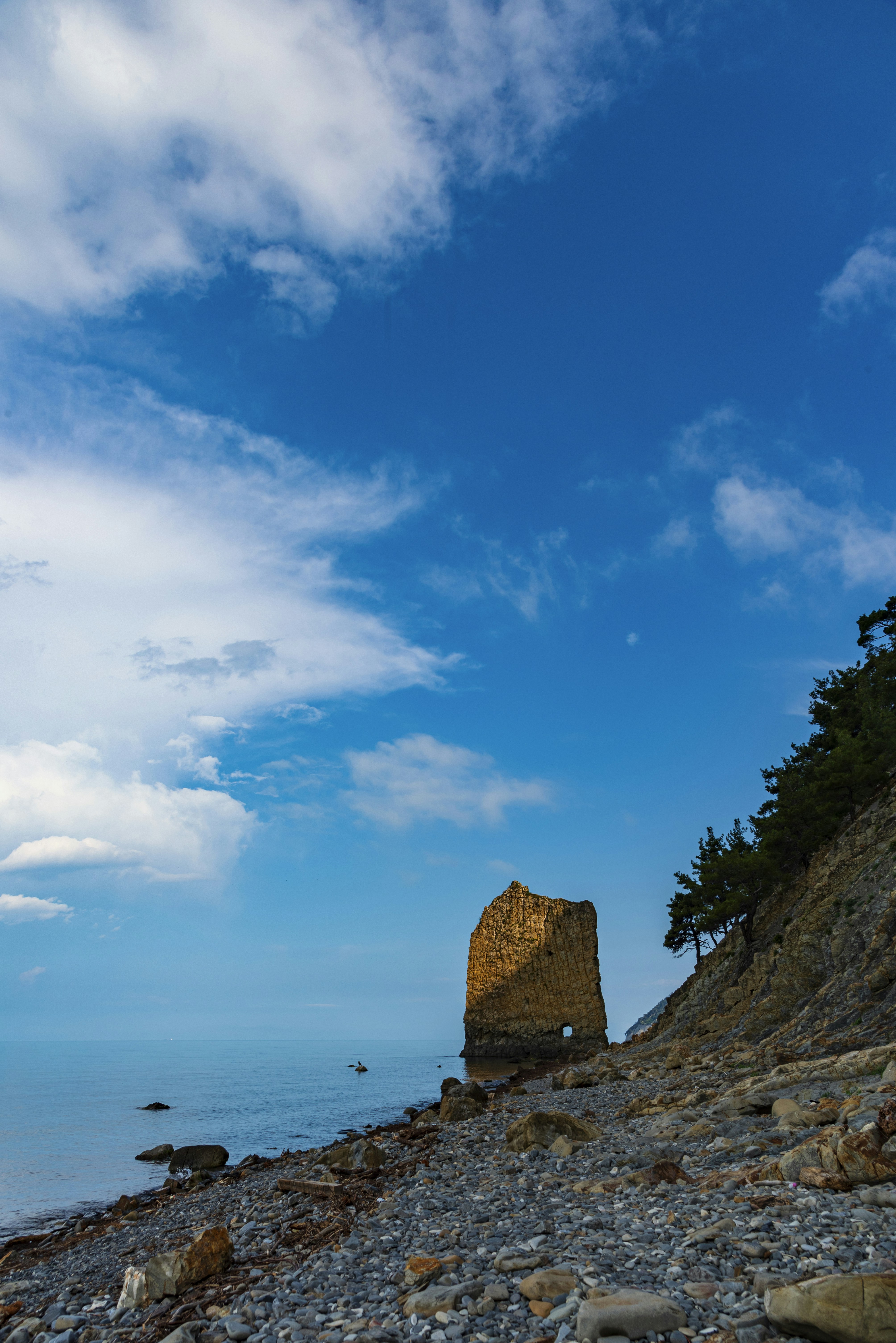A weathered coastal rock formation rises from a pebbled shoreline under a vivid blue sky.