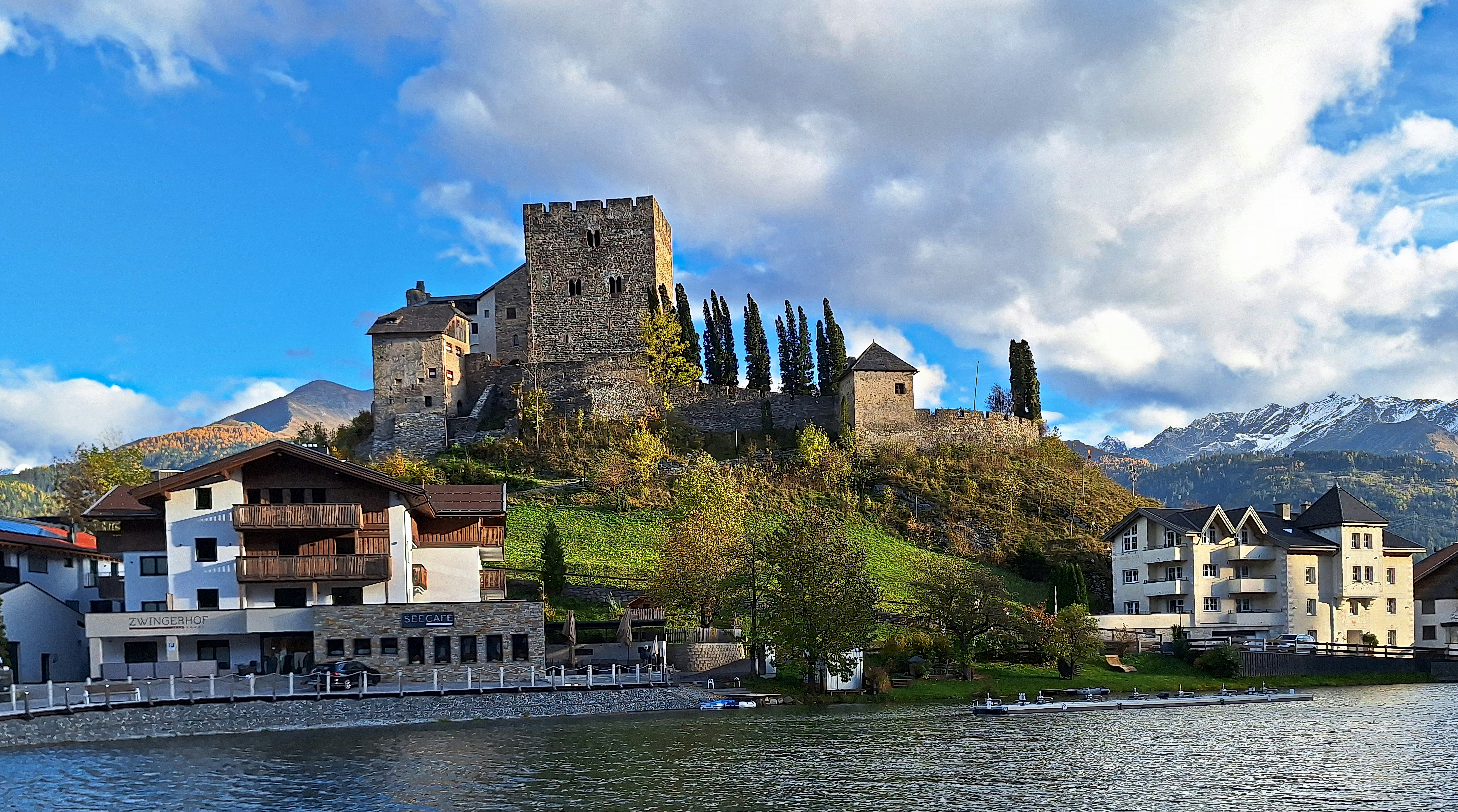 Historic castle overlook above a lakeside town with mountains in the distance; a tranquil riverside scene featuring blue sky and boats along the shore.