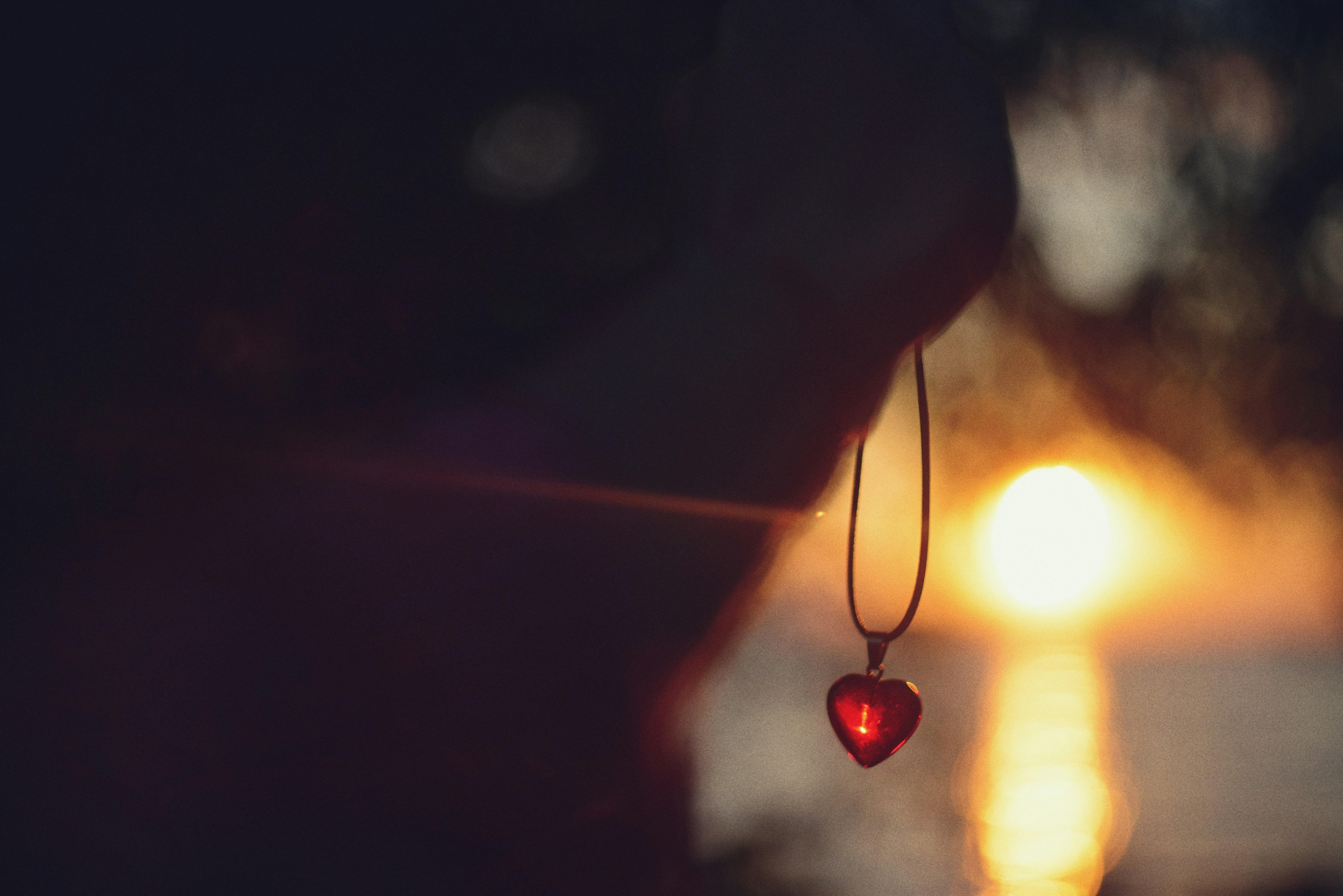 A dark silhouette of a person with a heart-shaped pendant hanging in the foreground, golden sunset reflecting on the water in the background.
