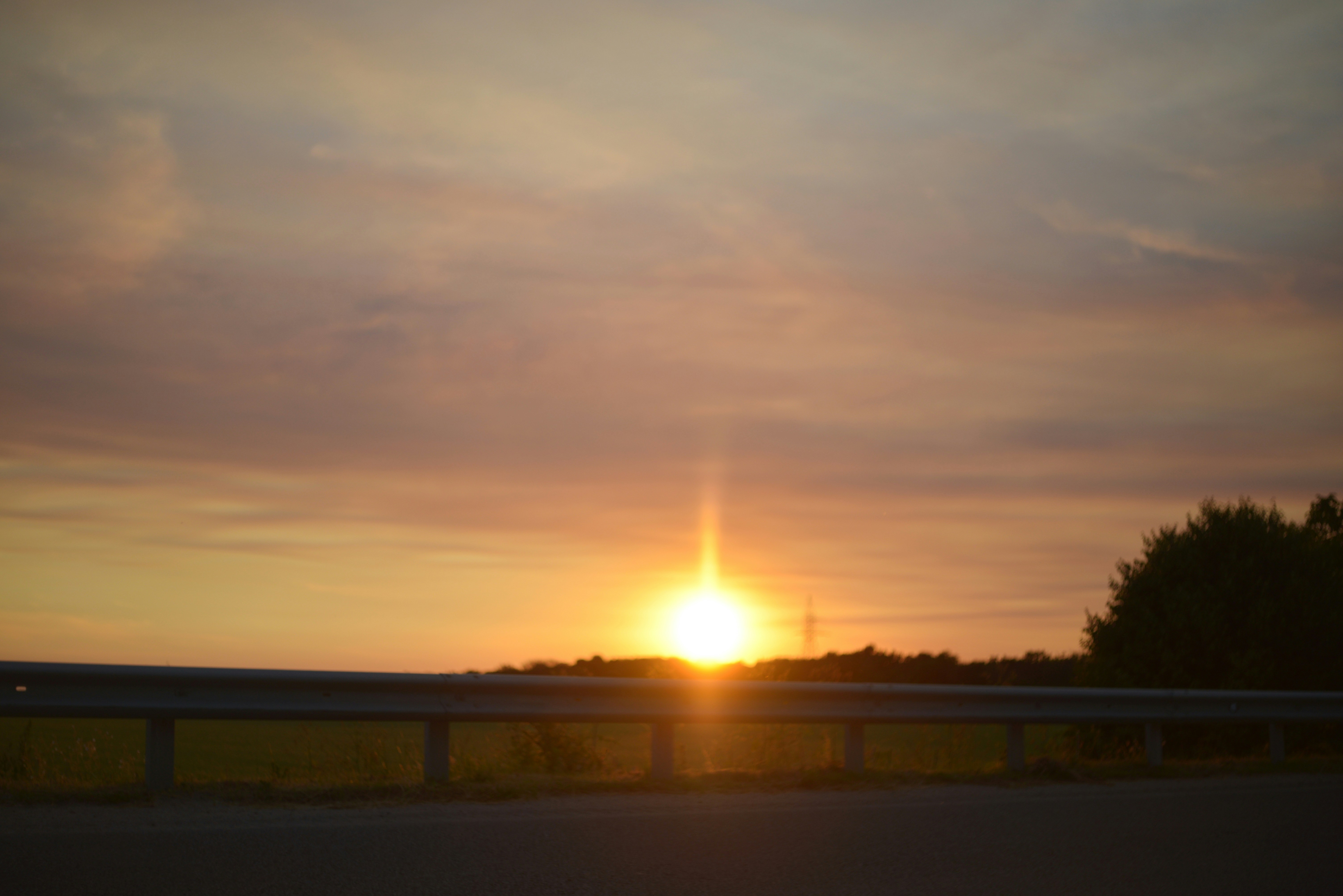 Warm sunset glow over a roadside scene with a low sun just above the horizon and a guard rail cutting across the foreground.
