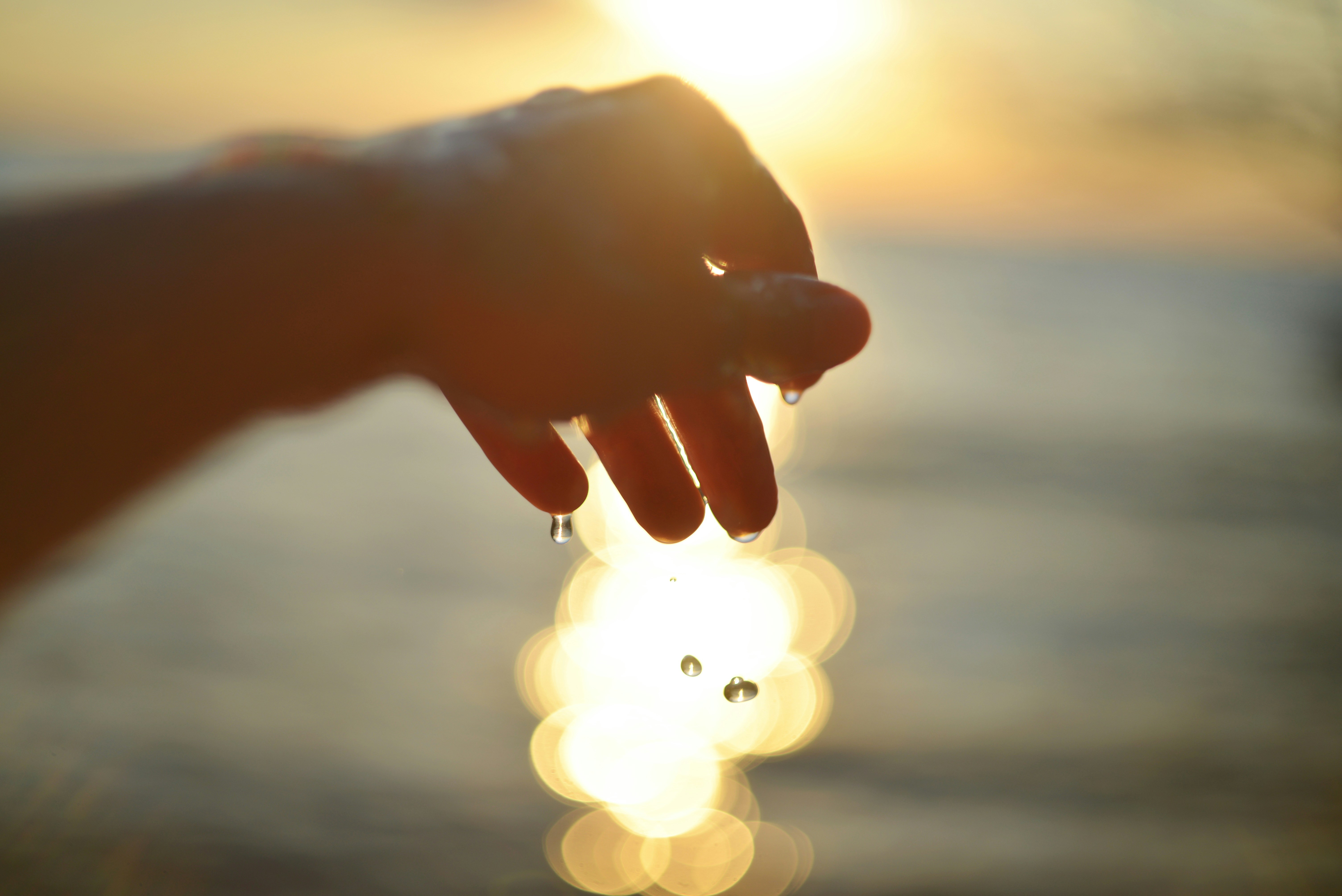 Close-up of a hand releasing droplets over a sunlit, bokeh-filled background; minimal foreground detail with warm, golden tones.