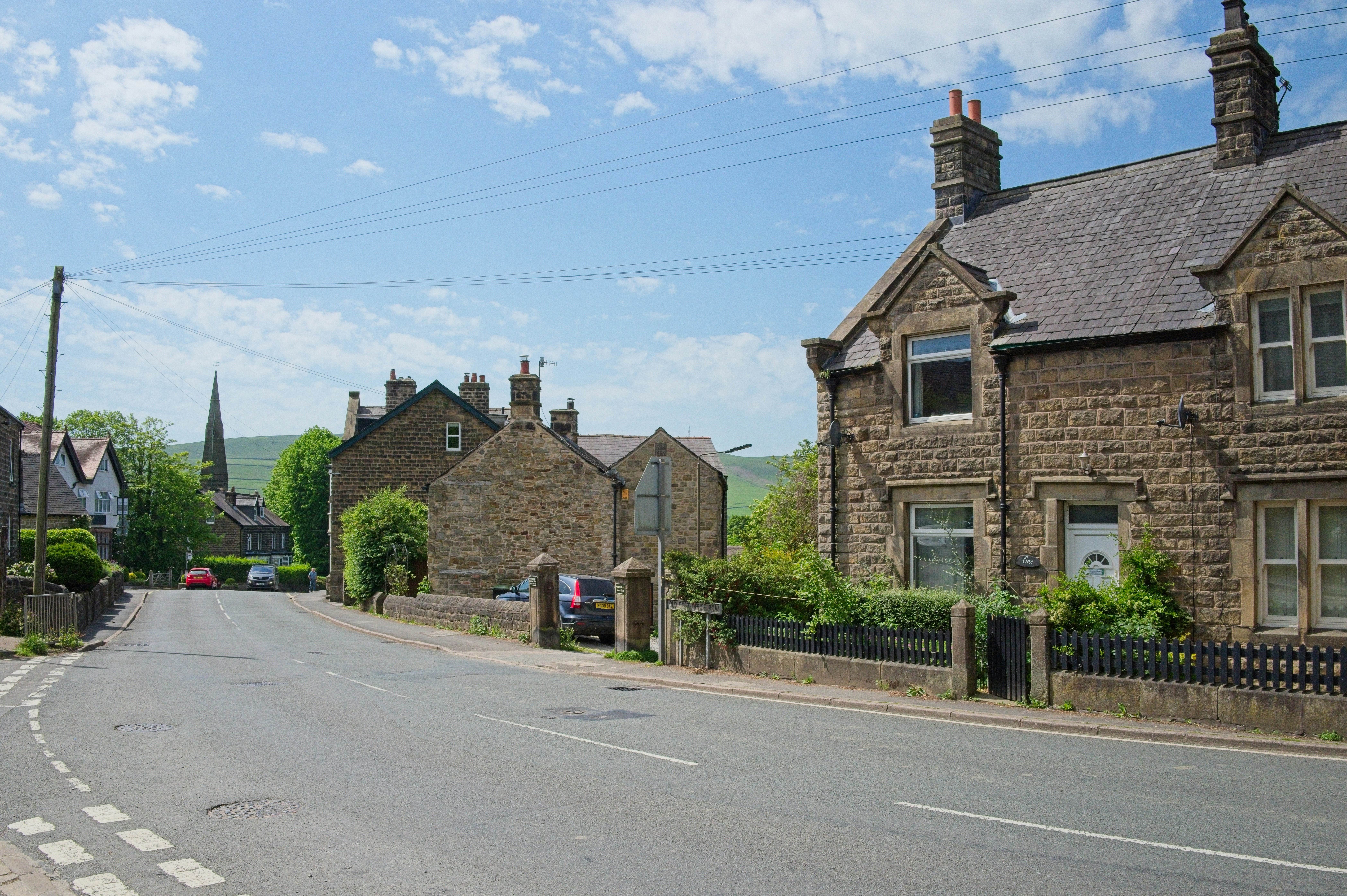 Sunlit village street with stone houses and parked cars along a quiet road, capturing the quaint English countryside ambiance.