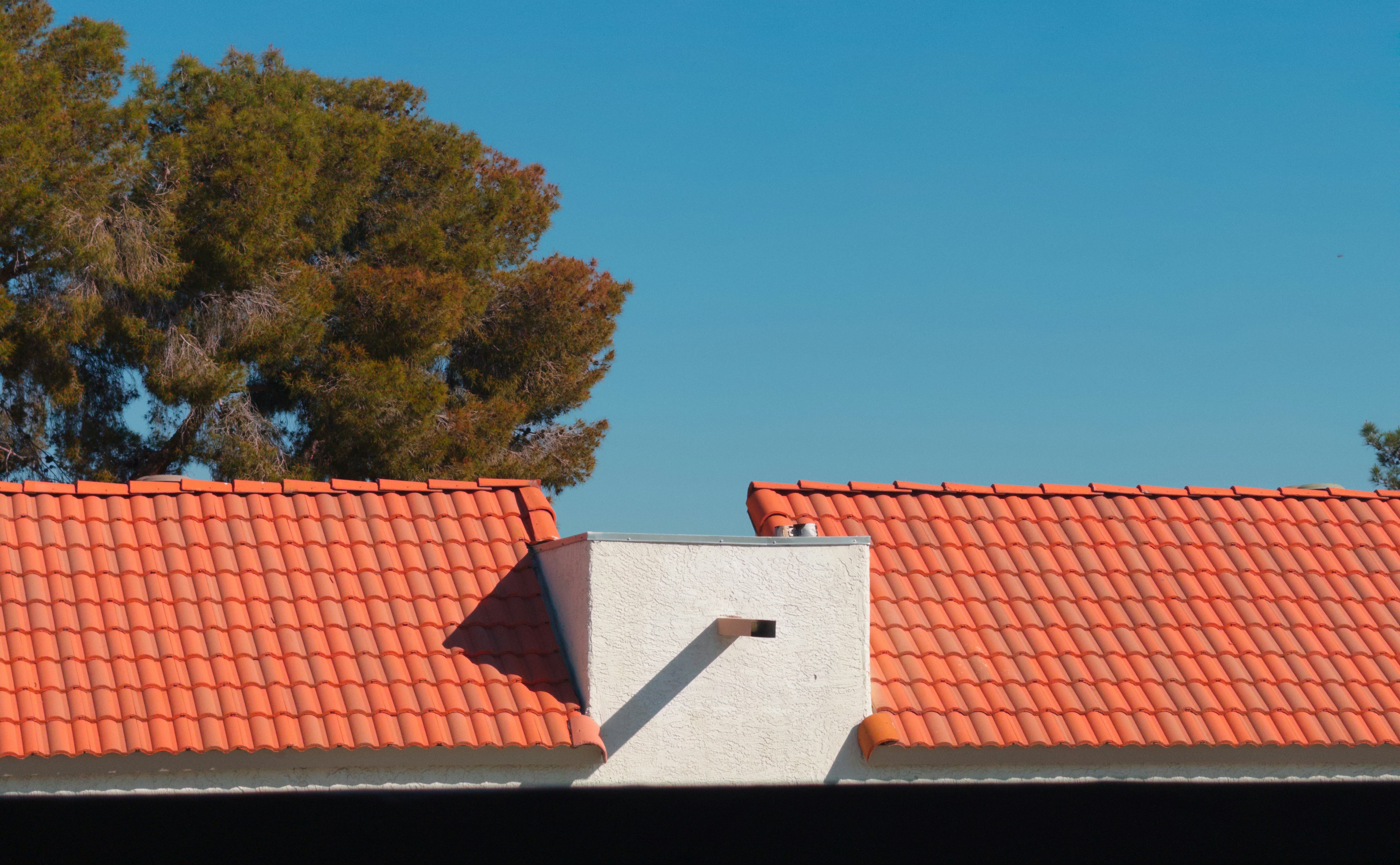 Clean architectural shot of a roof with red clay tiles against a bright blue sky, featuring a white chimney structure as the focal point.