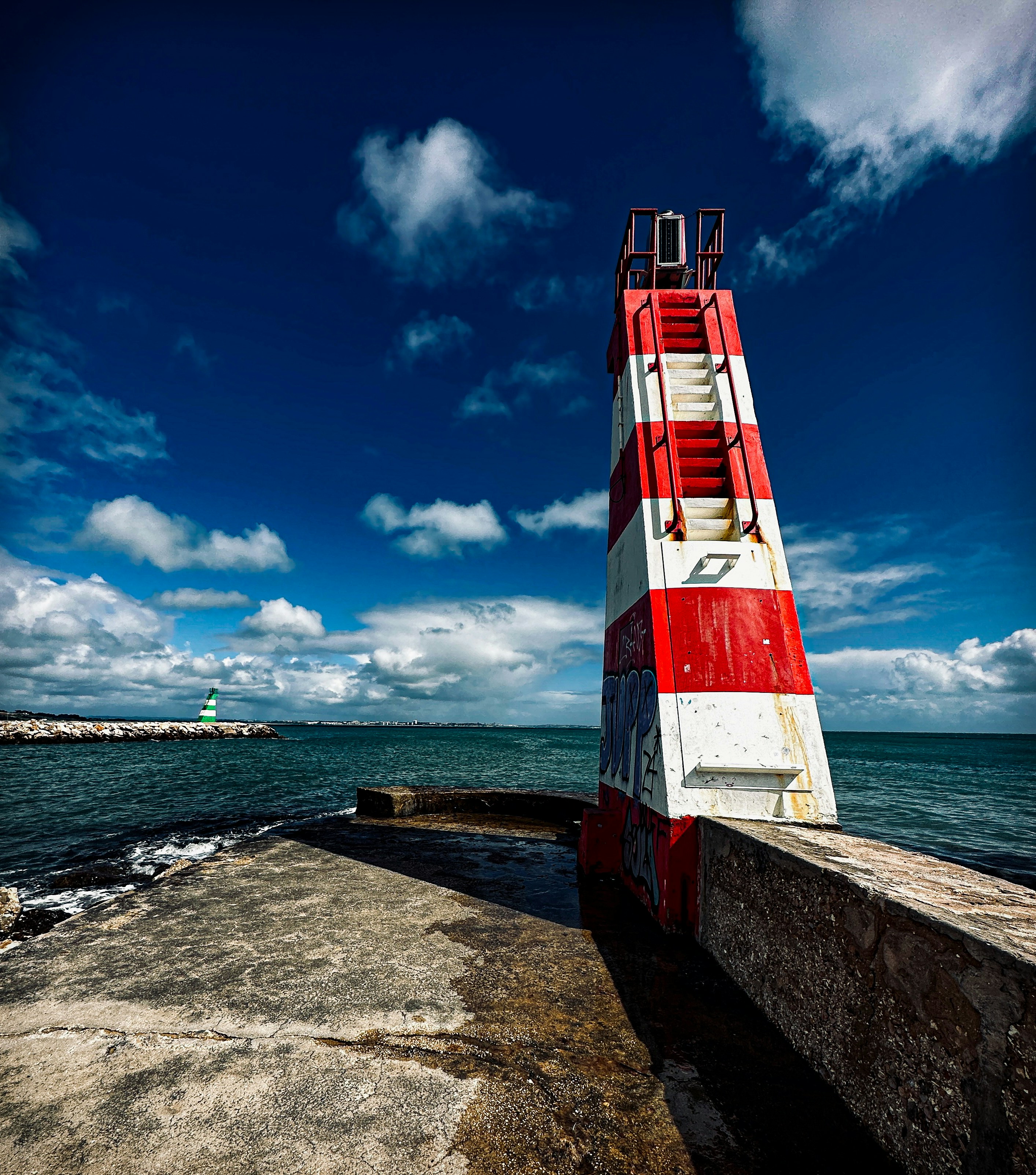 A bold striped lighthouse-like structure stands on a concrete jetty by the sea, with a deep blue sky and scattered clouds overhead.