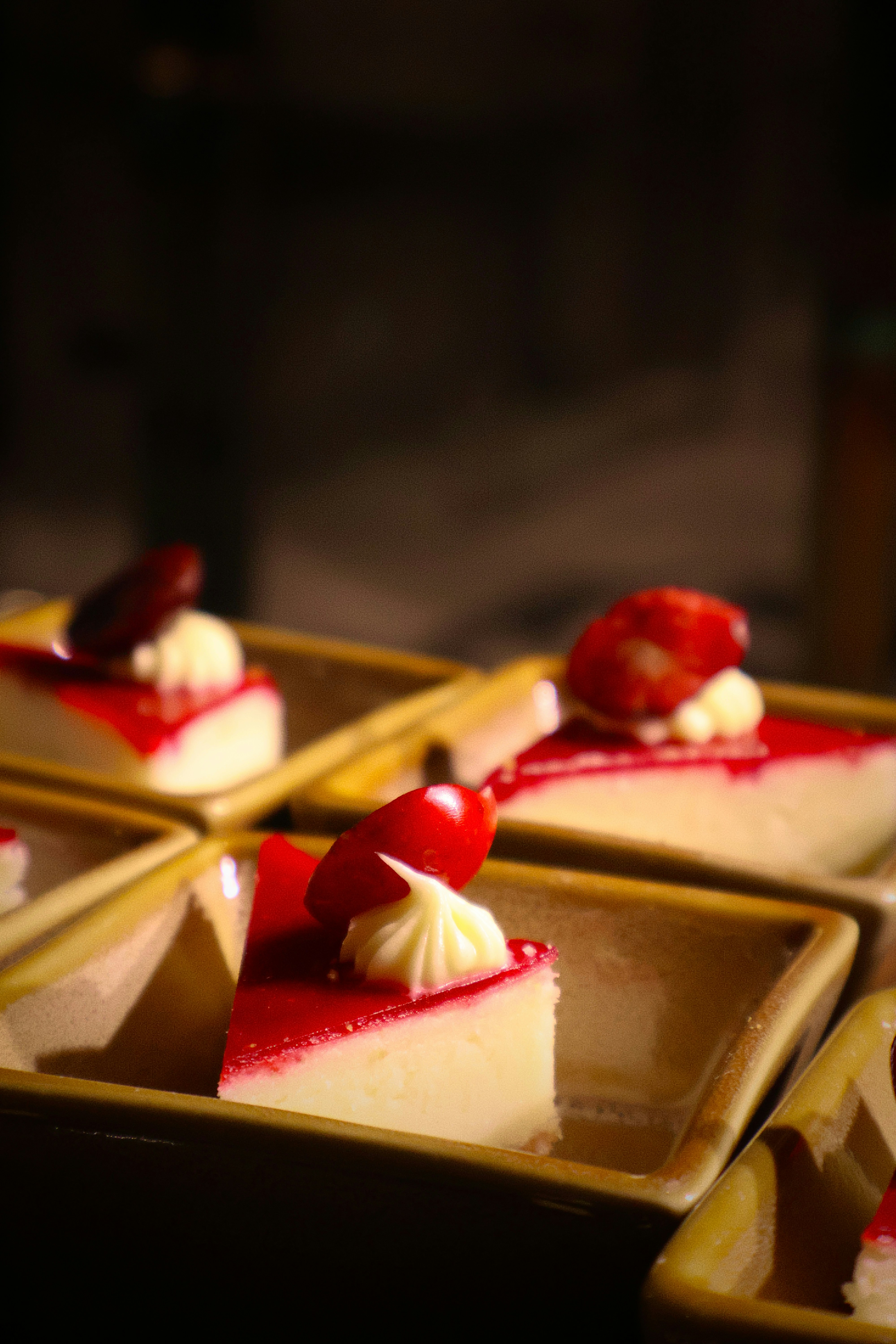 Creamy cheesecake squares topped with red glaze and whipped cream, arranged in golden serving trays. A dessert display focusing on individual portions.