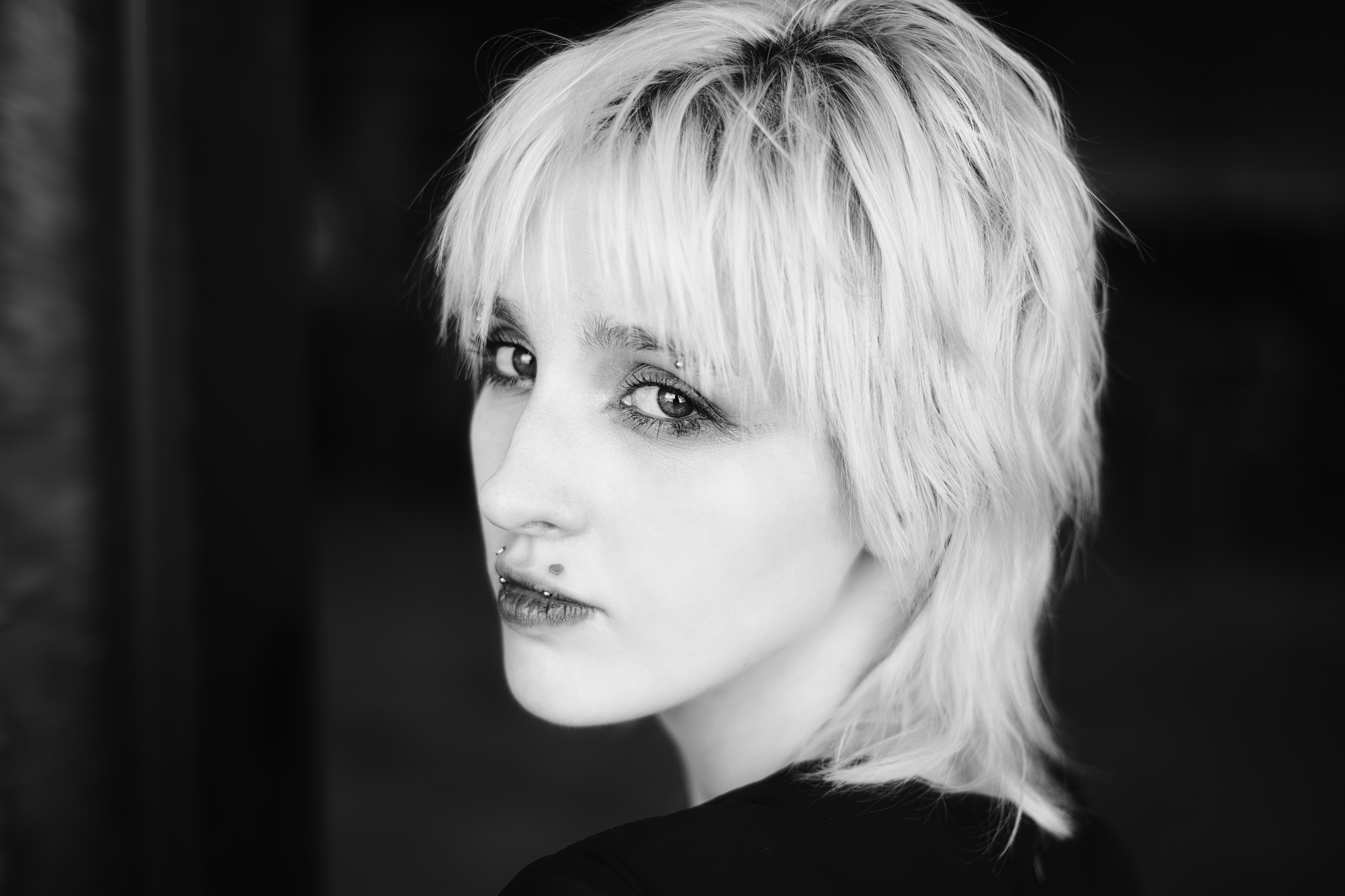 Monochrome portrait of a young woman with bleached hair, direct gaze, and subtle lip piercing; the frame emphasizes her face against a dark background.