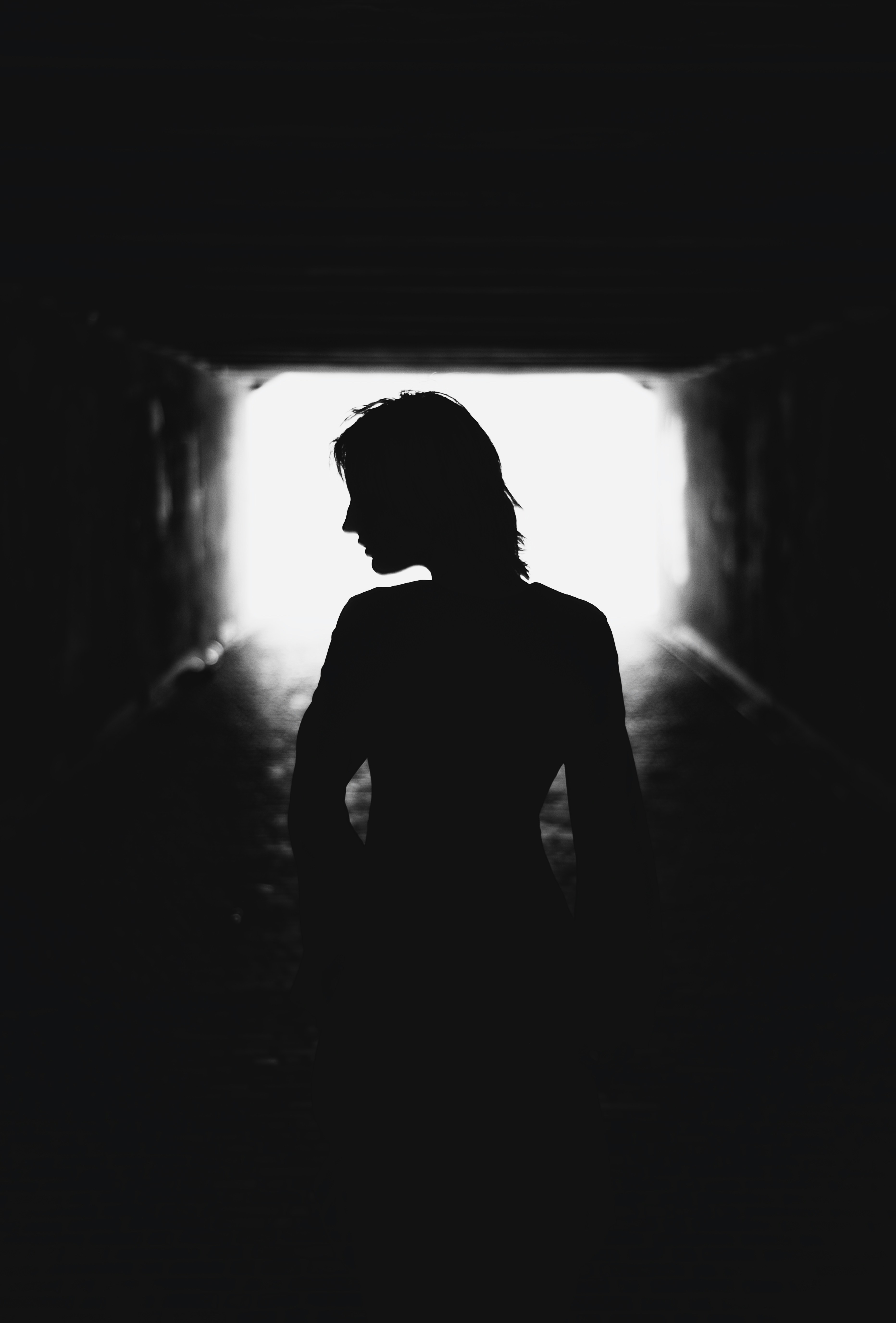 Silhouette of a person standing in a dark tunnel with bright light behind, emphasizing the outline and negative space. The image is a high-contrast black-and-white portrayal, focusing on form over detail.