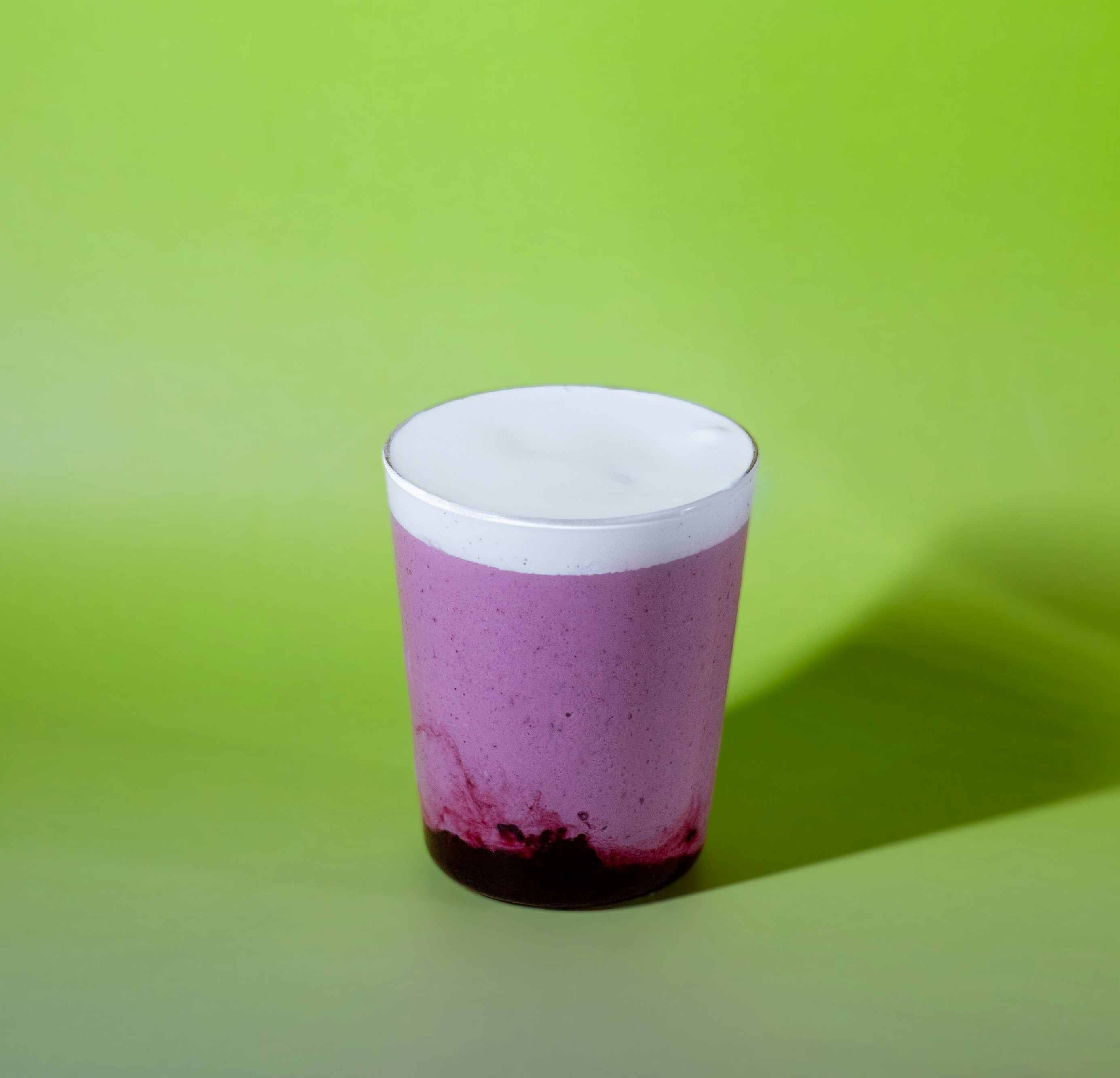 A pink layered beverage in a clear glass sits on a lime-green surface with a soft shadow, framed to emphasize its height and color gradient. This is a photograph.