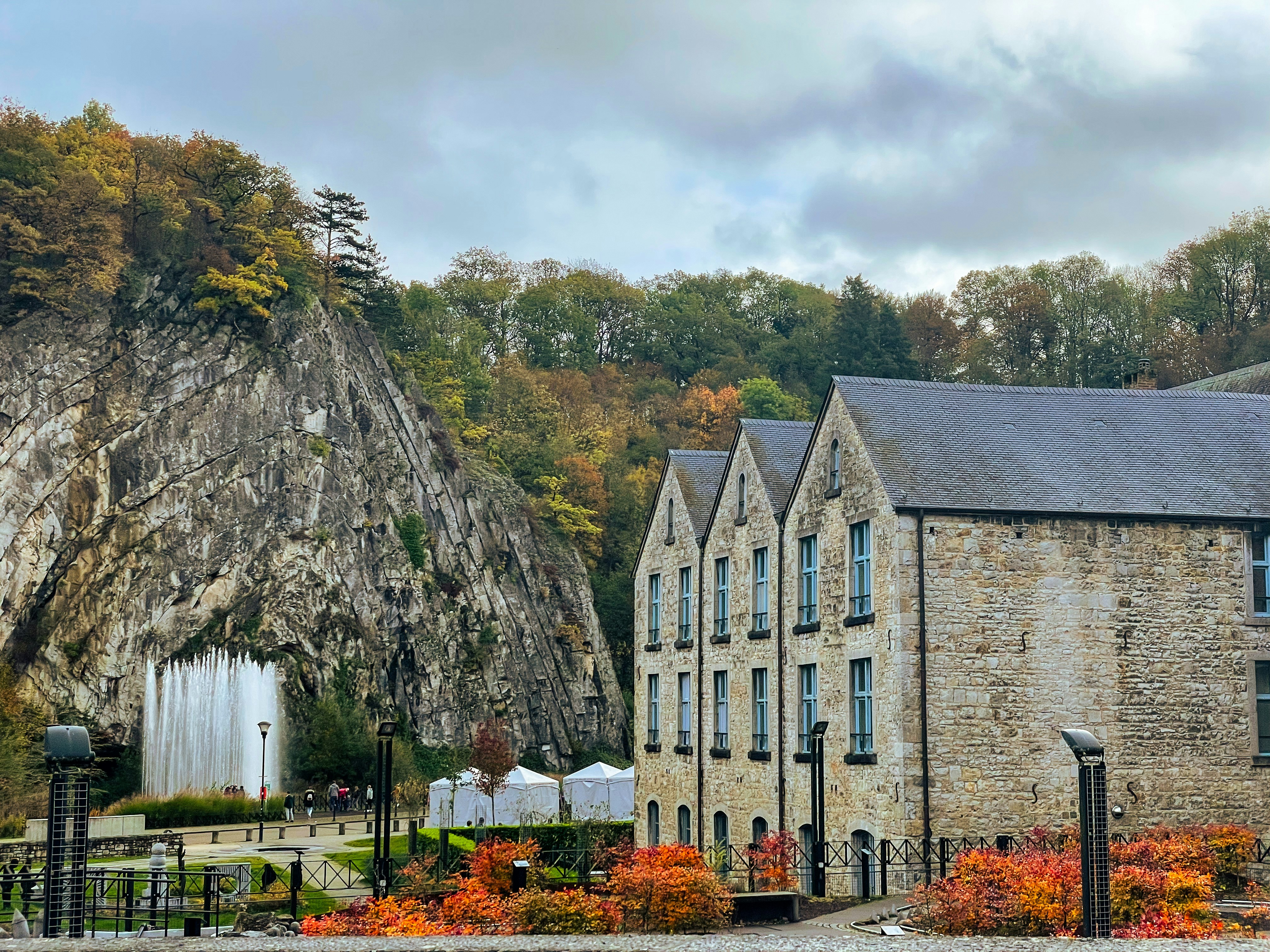 Stone mill buildings sit at the base of a rocky cliff, with fall foliage and a small fountain in the background.