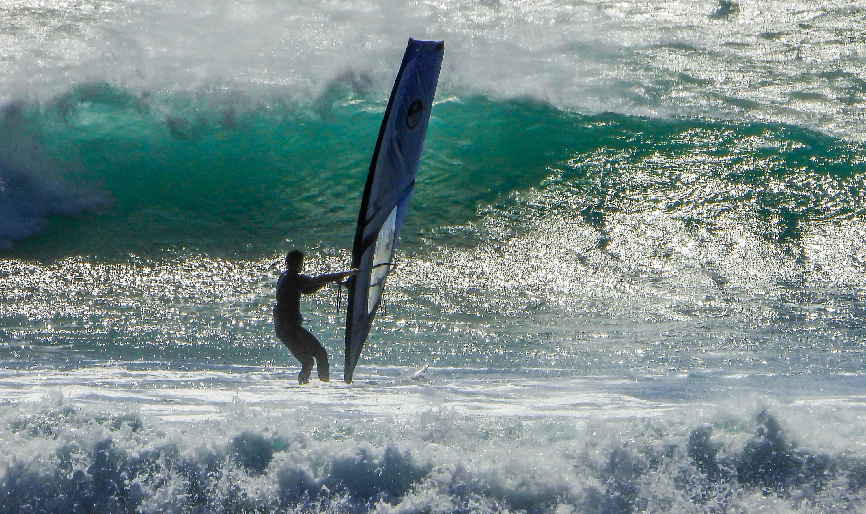 Windsurfer glides on turquoise waves with a sail lifted, sunlight reflecting on the water’s surface.