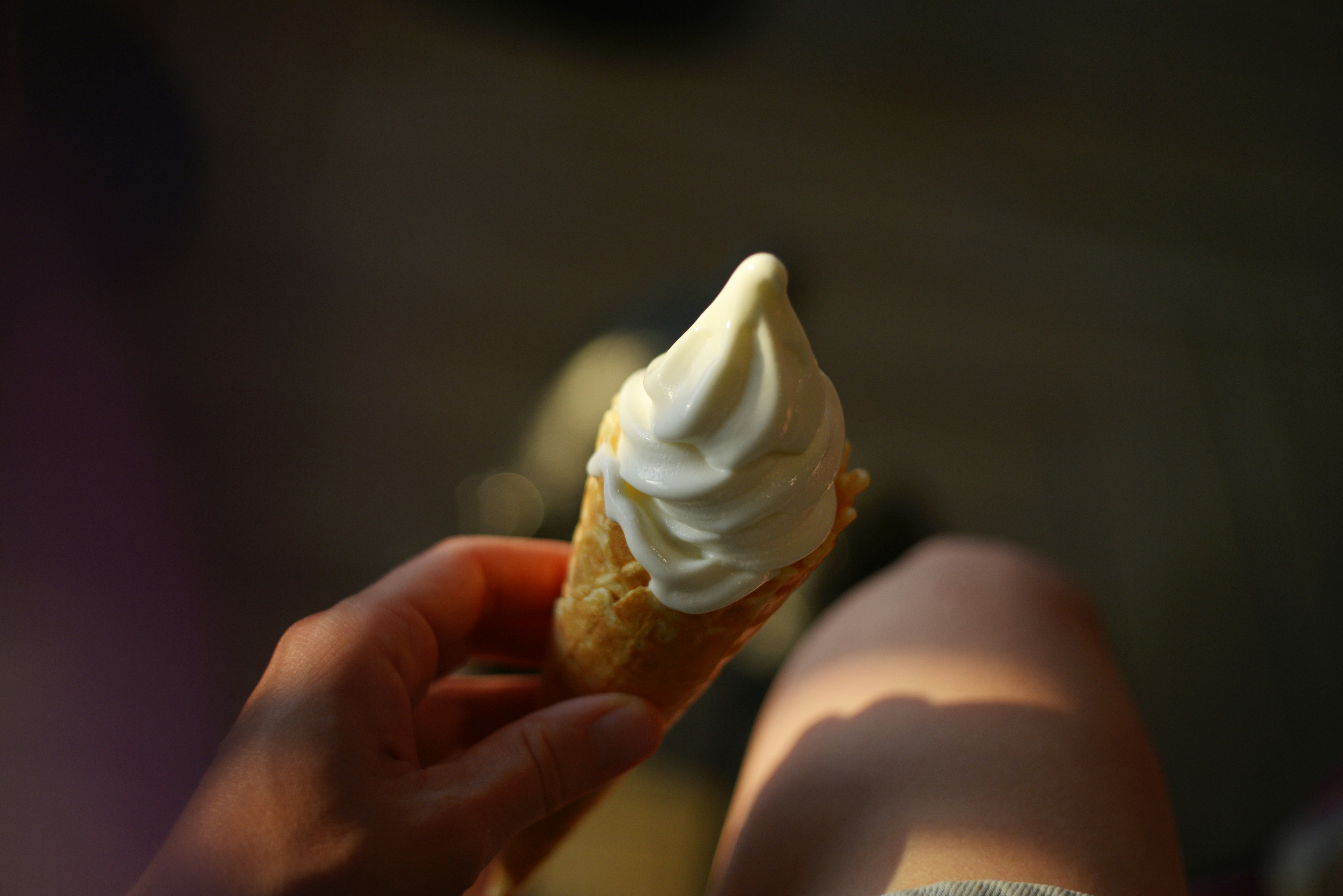 Warm-lit hand-held ice cream cone with shallow depth of field and blurred background, focusing on the creamy swirl and the texture of the waffle cone.