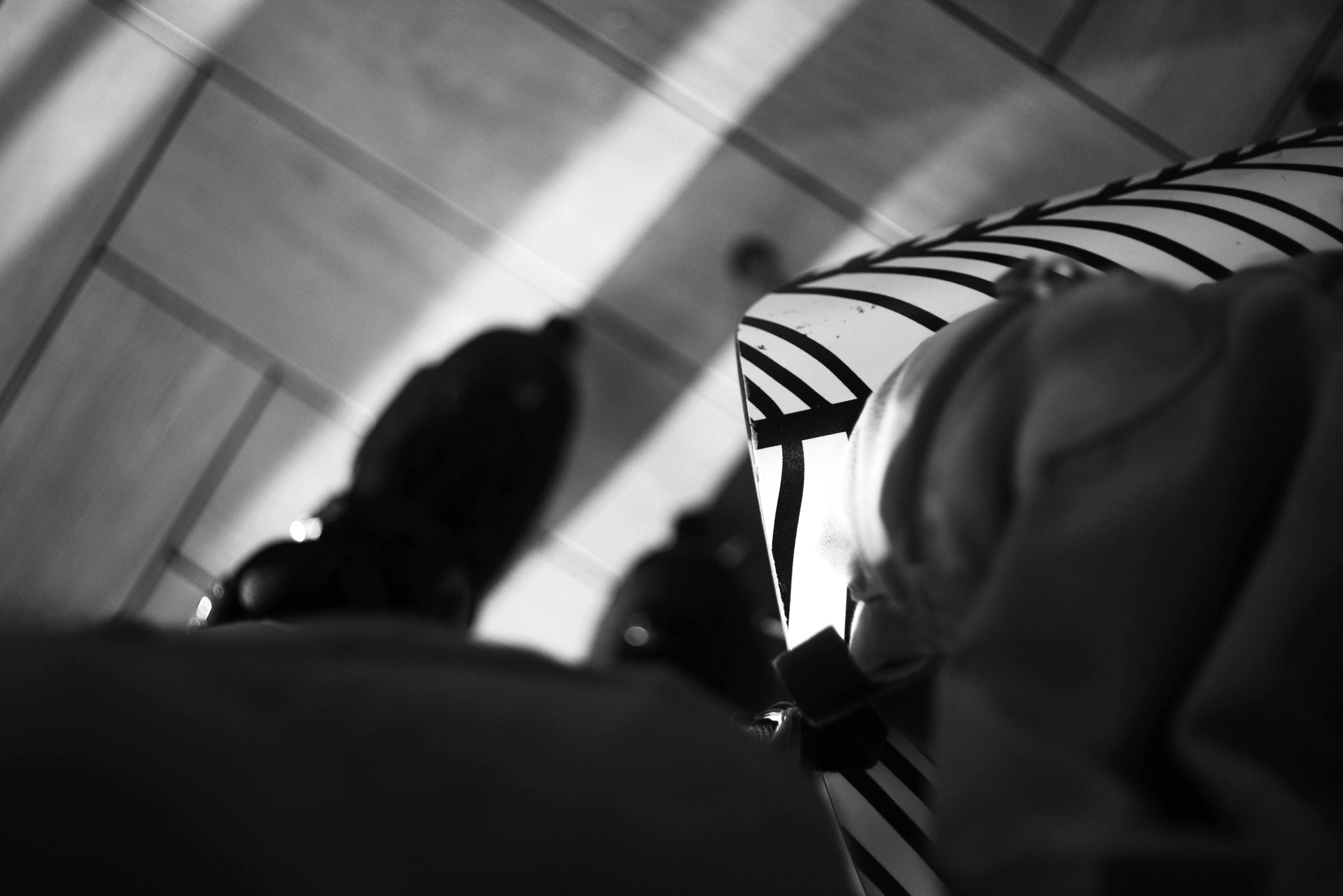 Monochrome scene of a person standing or walking in an indoor corridor, viewed from above, with patterned seating and shoes at the bottom of the frame. The composition emphasizes perspective and texture, suggesting a candid moment in a waiting area.