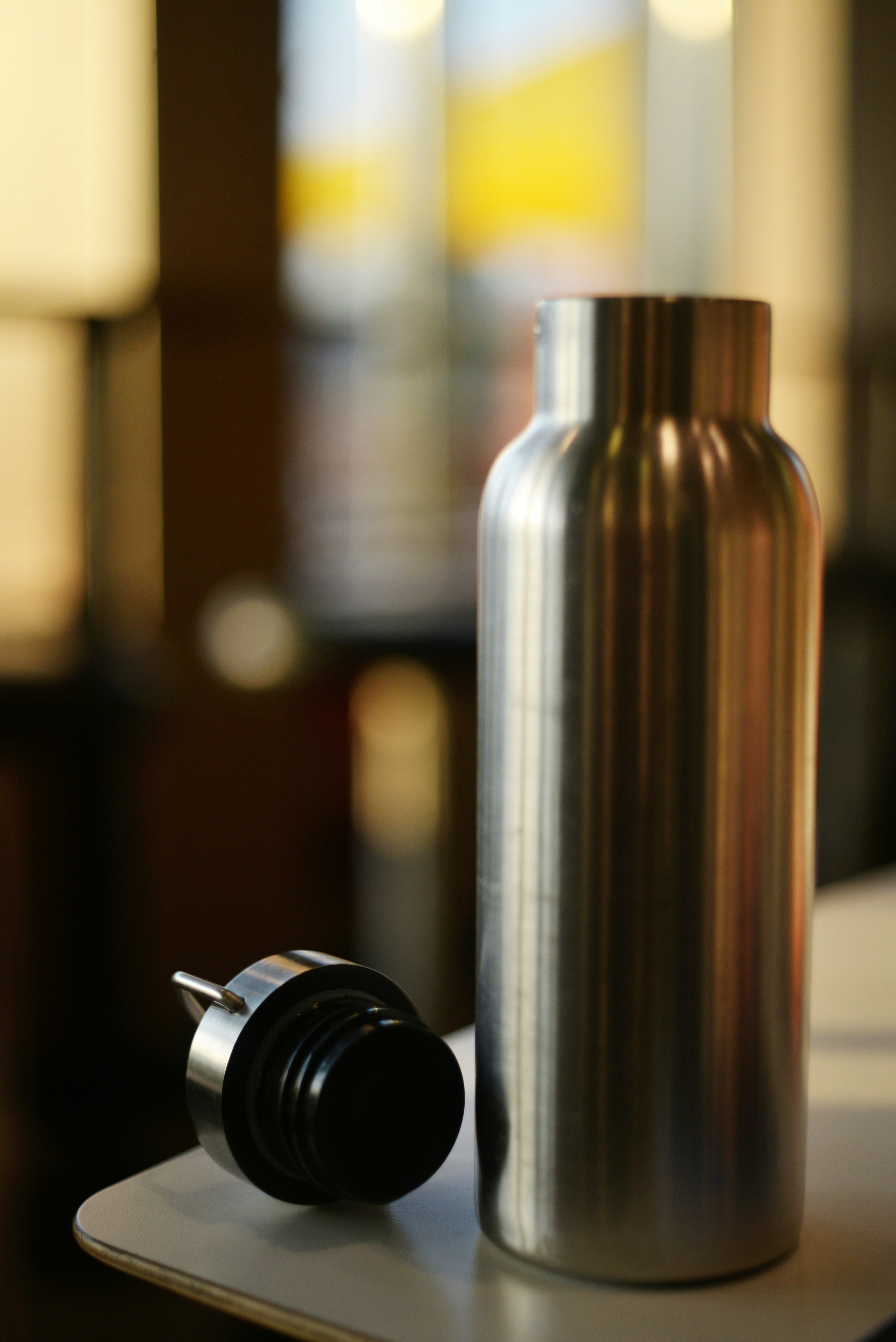 Close-up of a metallic bottle and detached cap resting on a desk, with a blurred background suggesting a workshop or kitchen setting.