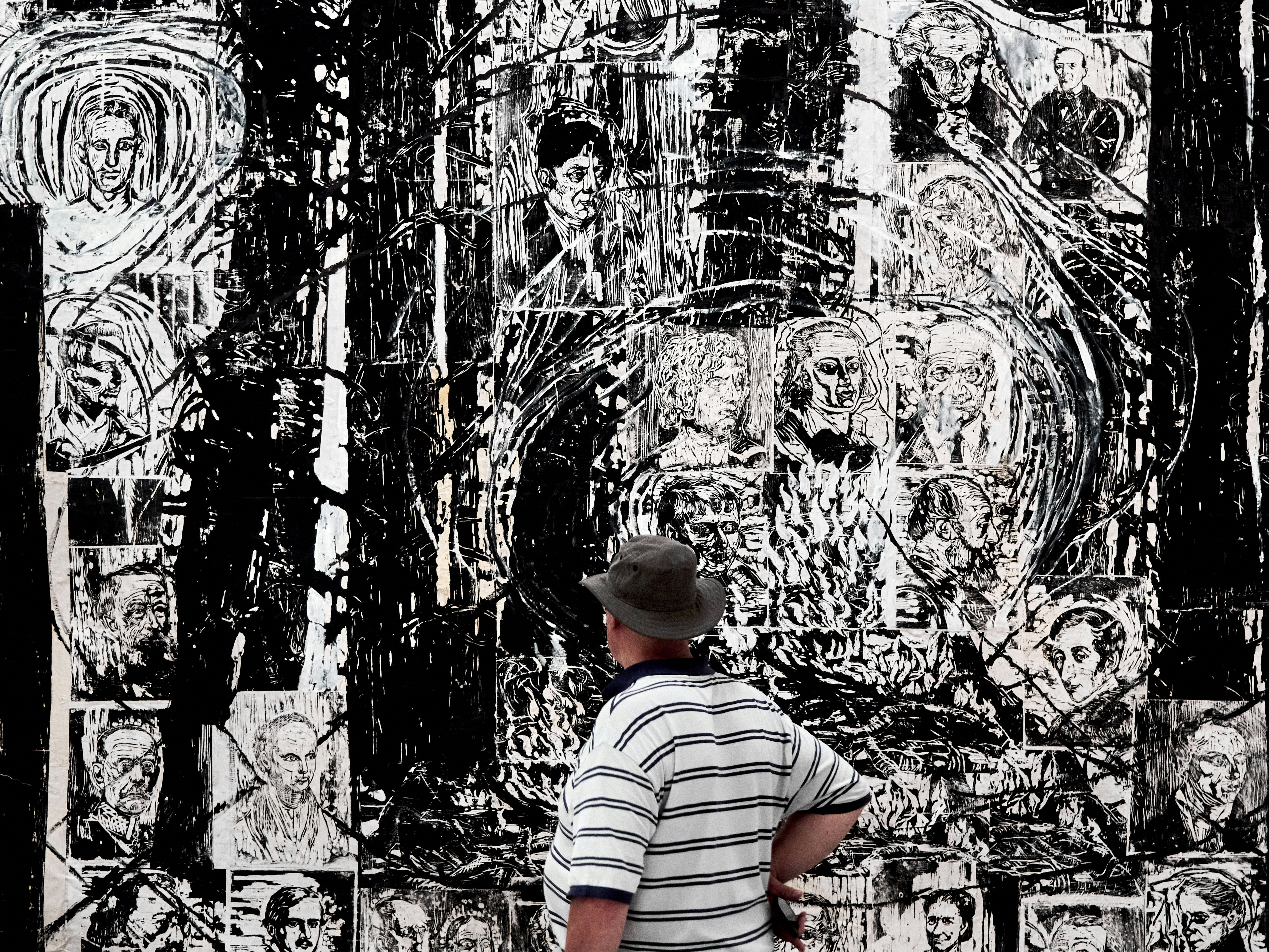 A man wearing a striped shirt and hat stands with hands on hips, facing a dense wall covered in black-and-white portraits and abstract drawings that resemble a mural or collage.