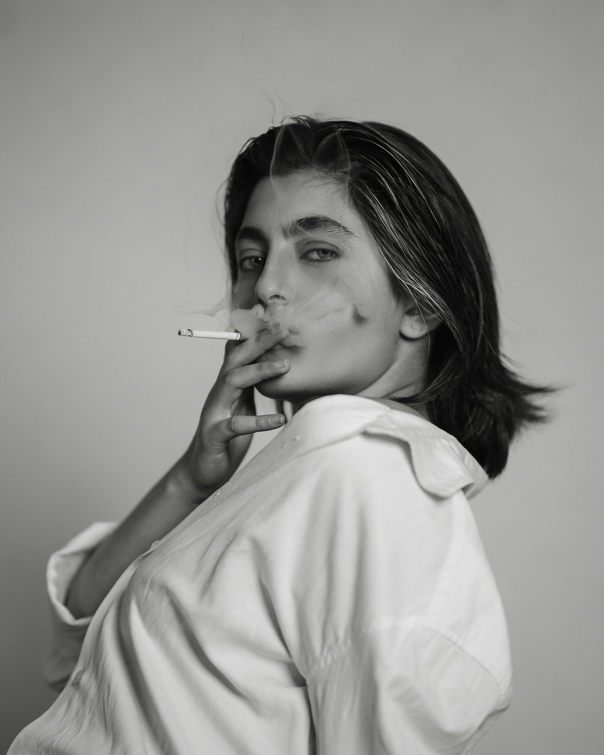 Monochrome portrait of a woman smoking a cigarette, head turned over her shoulder, wearing a light shirt; the image emphasizes mood and texture with soft lighting.