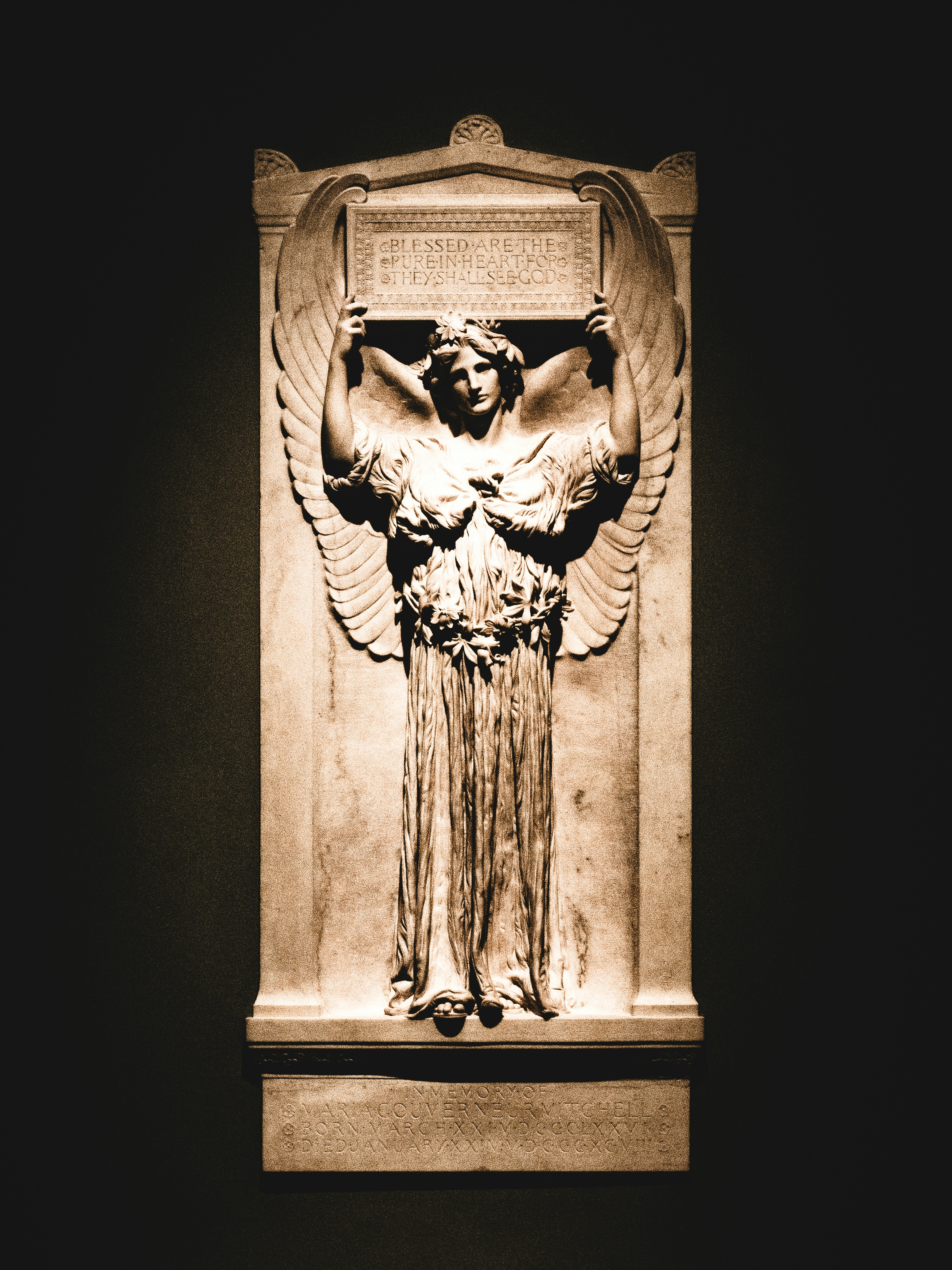 A carved angel relief holding a plaque, illuminated against a dark background, emphasizing the statue's wings and flowing robes. The sculpture appears as a standalone monument, captured with warm, cinematic lighting.