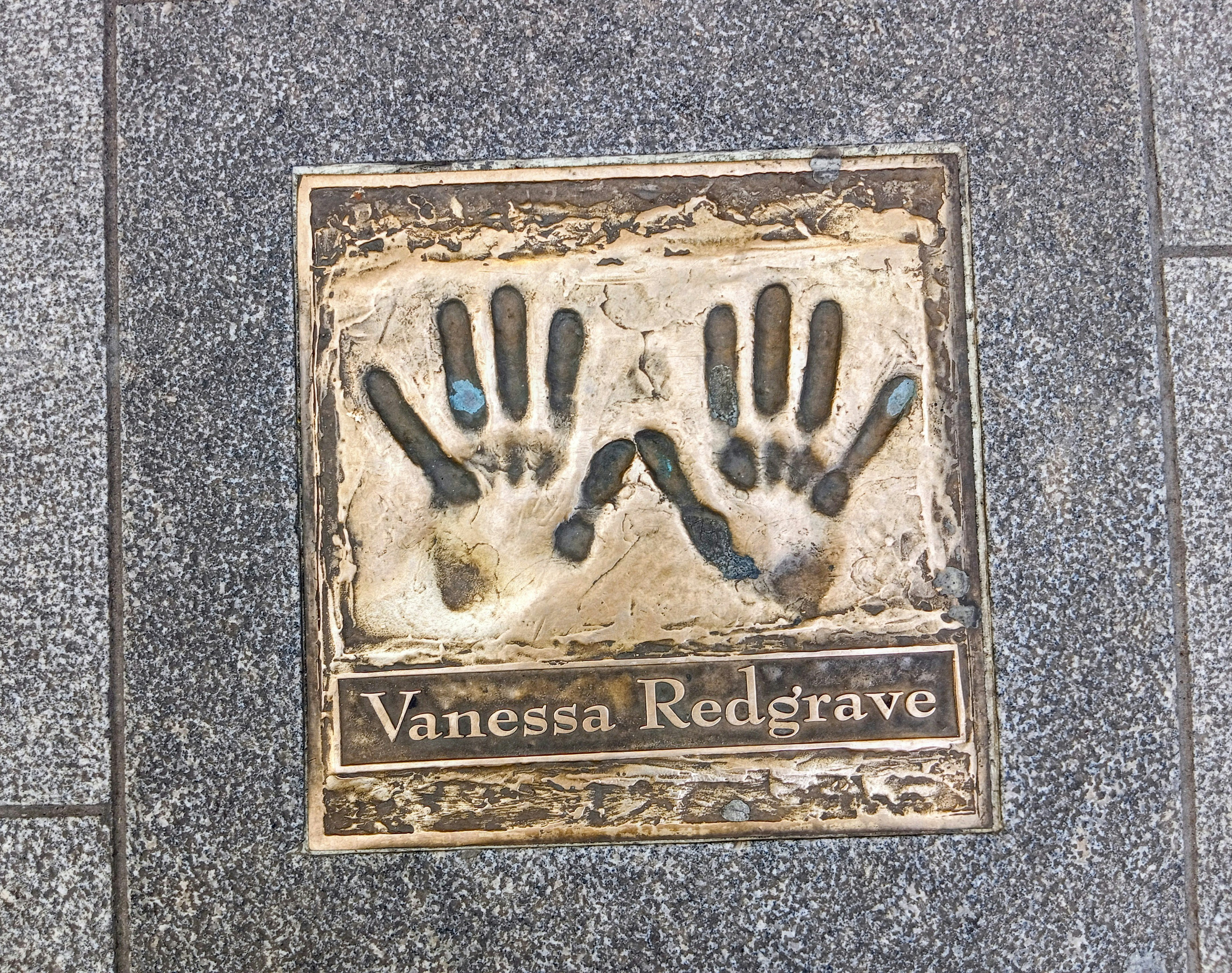 Bronze plaque with raised handprints and nameplate embedded in pavement; commemorative tribute on a sidewalk.