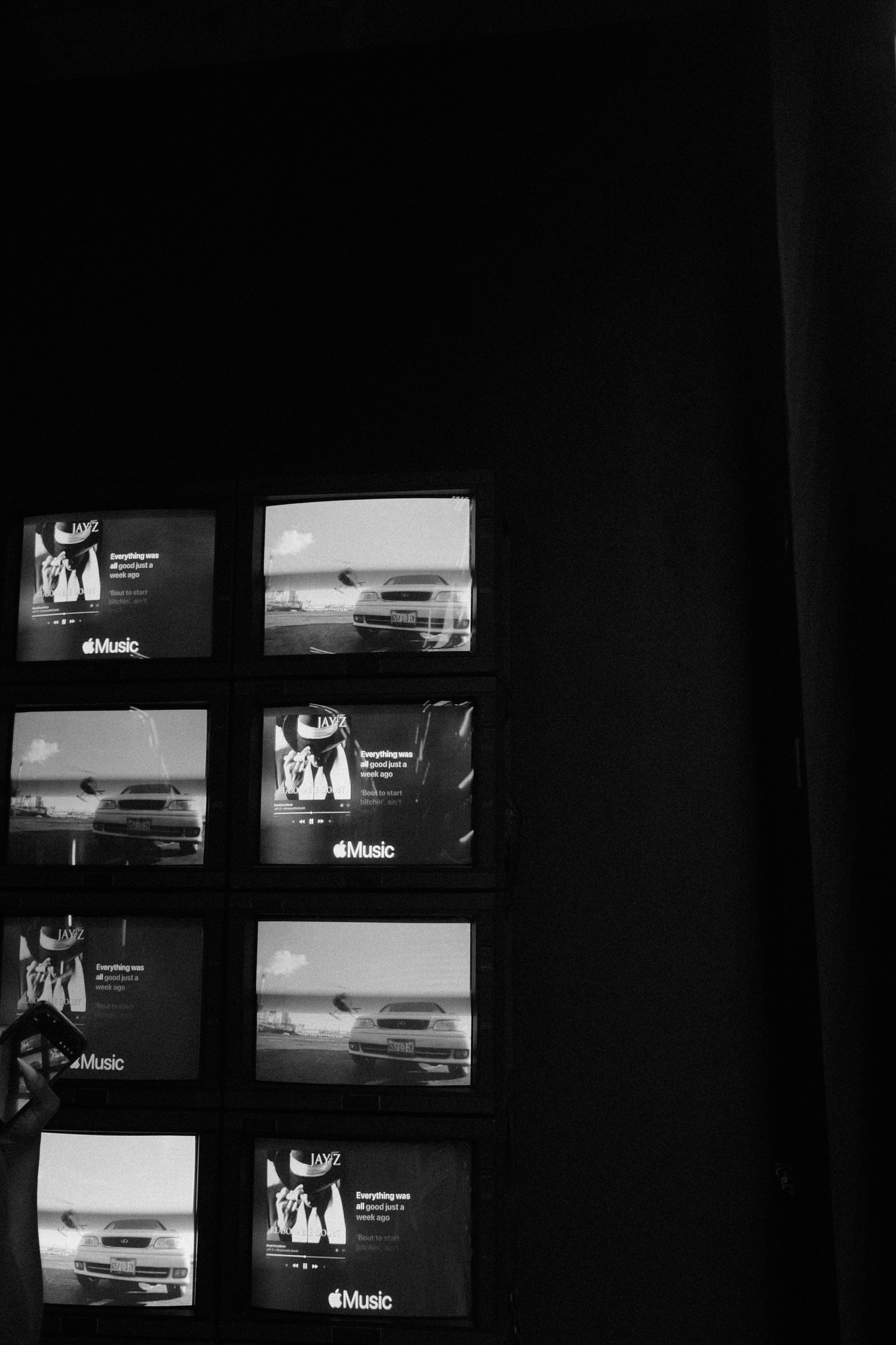 Monochrome wall of small screens displaying car images and music ads, forming a grid-like mosaic in a dark interior.