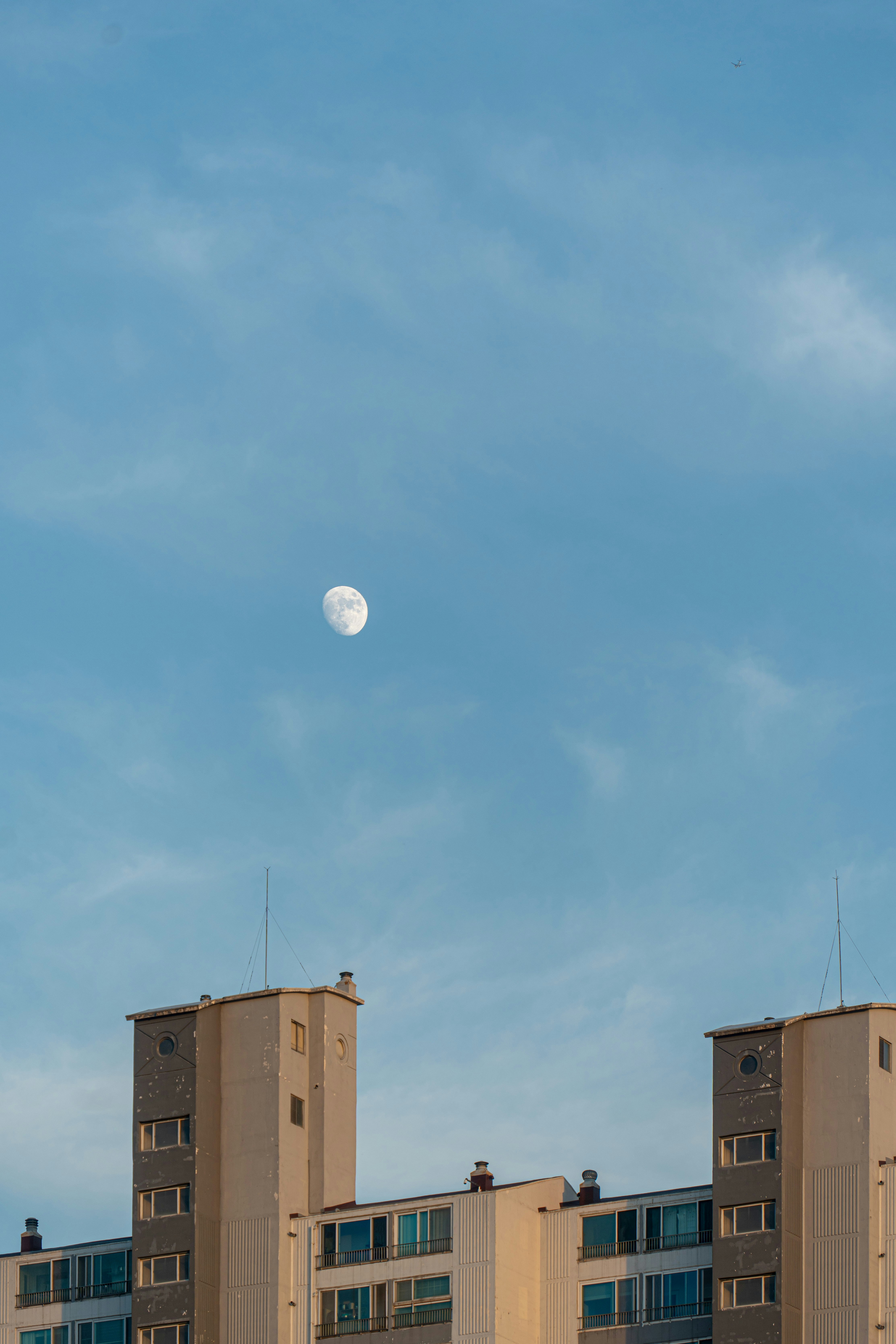 Soft blue sky with a rising moon over two modern apartment towers and a few rooftop vents. This image captures a tranquil urban moment and the geometry of the buildings against the open sky.