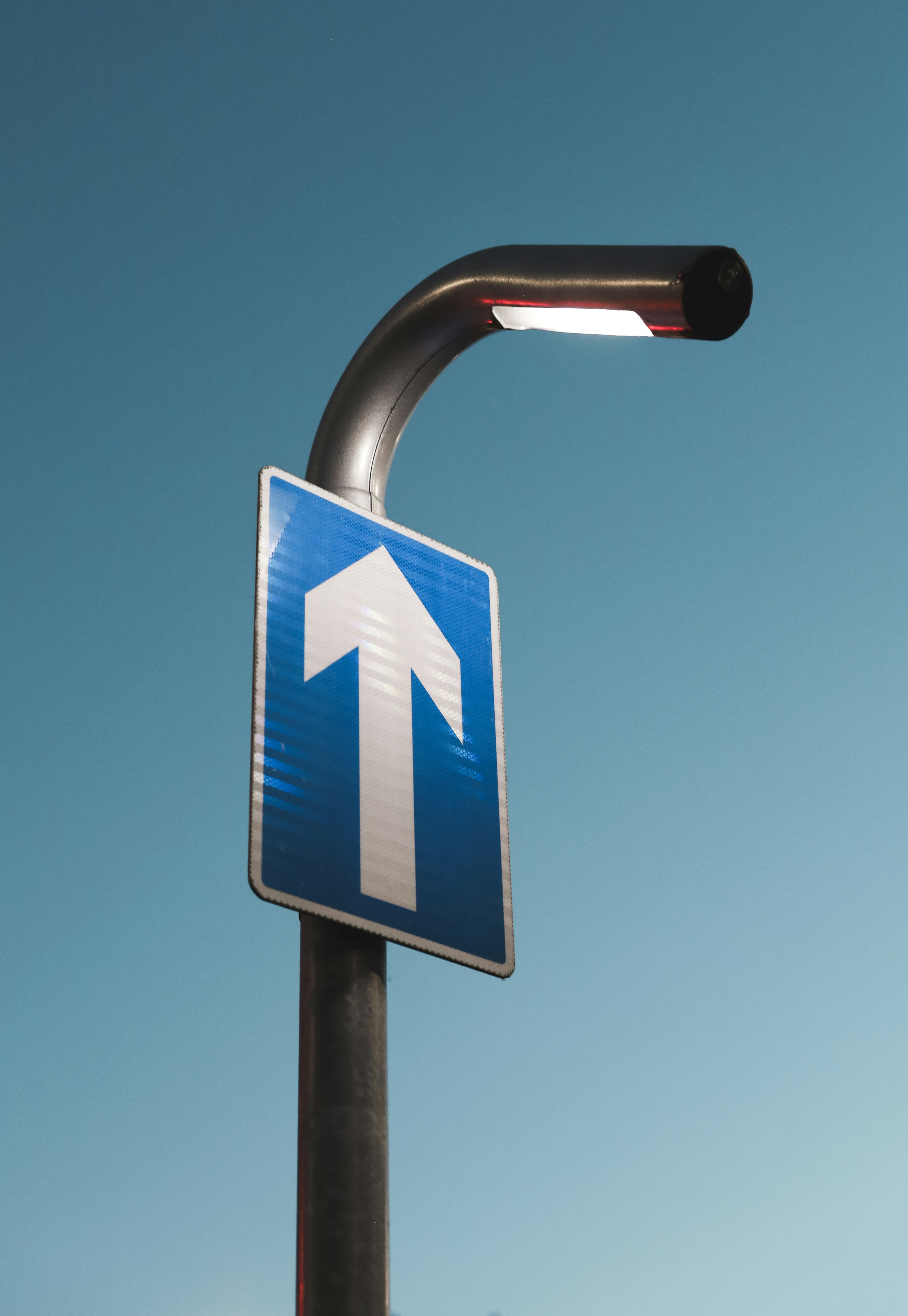 A blue upward-arrow sign atop a metal post against a clear blue sky, with a curved streetlight arch at the top; a simple urban street cue captured with clean contrast.