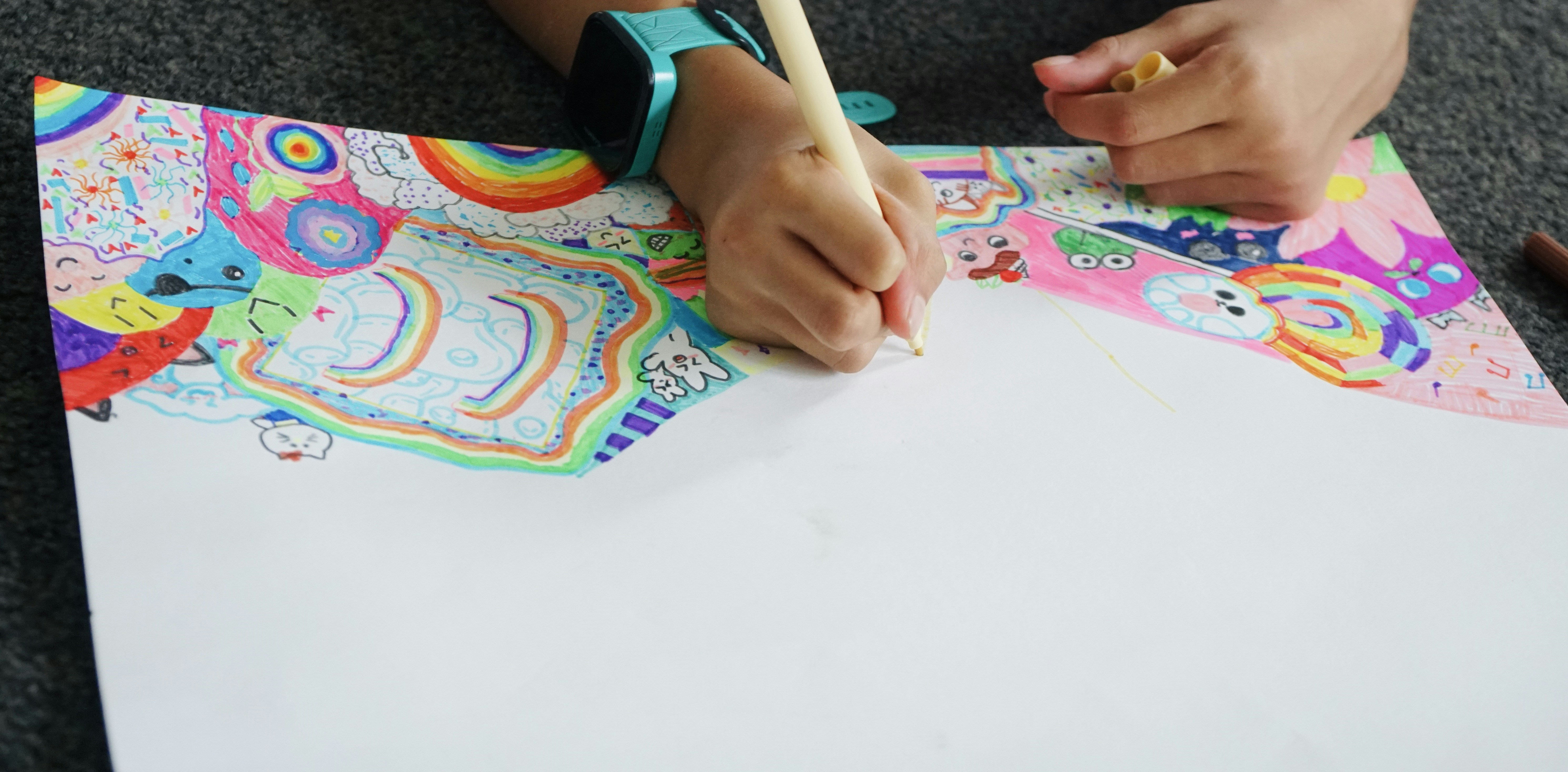 Bright crayons and a child's hand trace vivid patterns on a large sheet as colorful drawings spill toward the edges. A playful, artistic scene captured during a creative session.