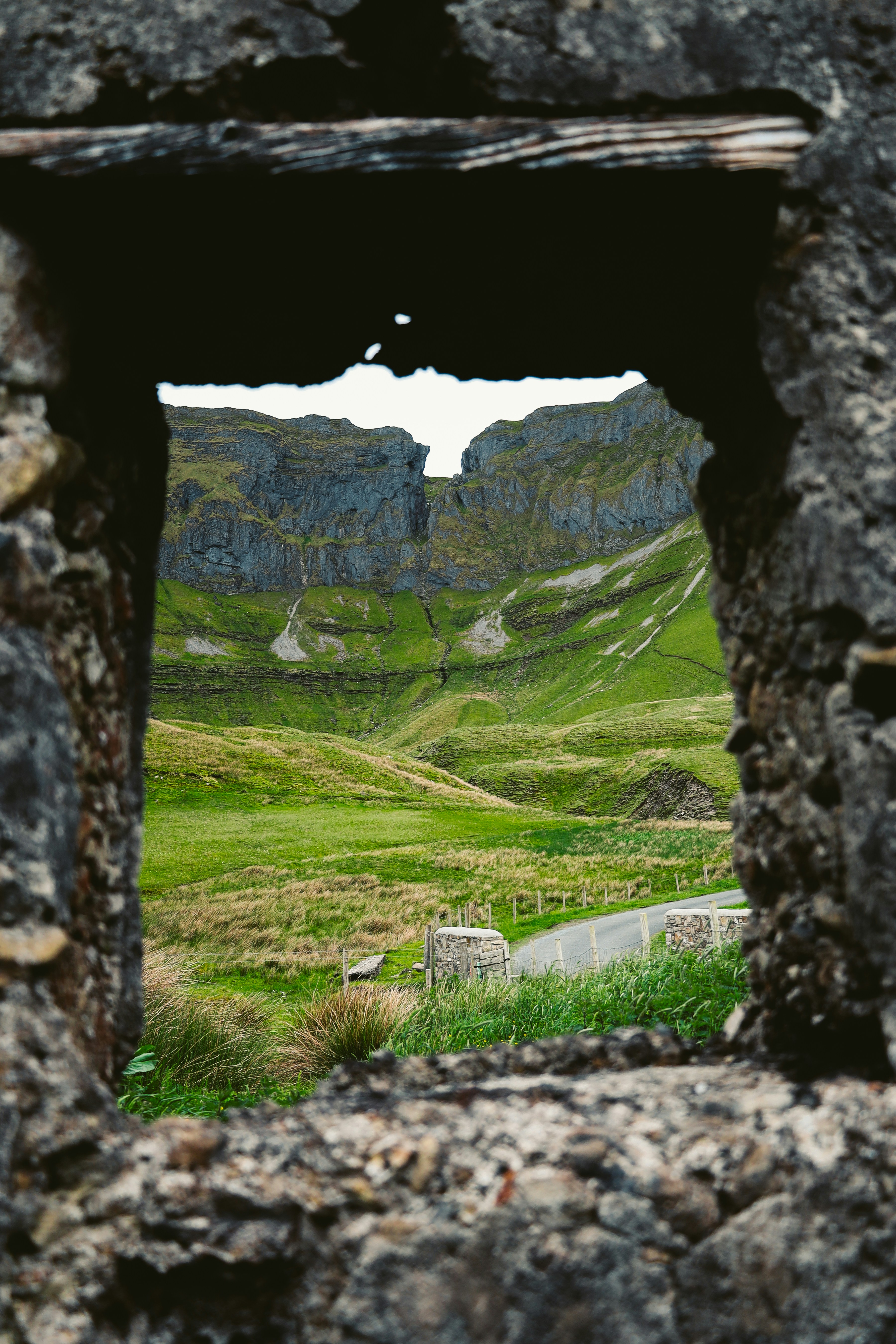 Lush green valley with rocky cliffs framed by a weathered window opening in stone, leading to a winding path and distant gate. A landscape shot emphasizing depth and contrast between foreground ruin and expansive countryside.