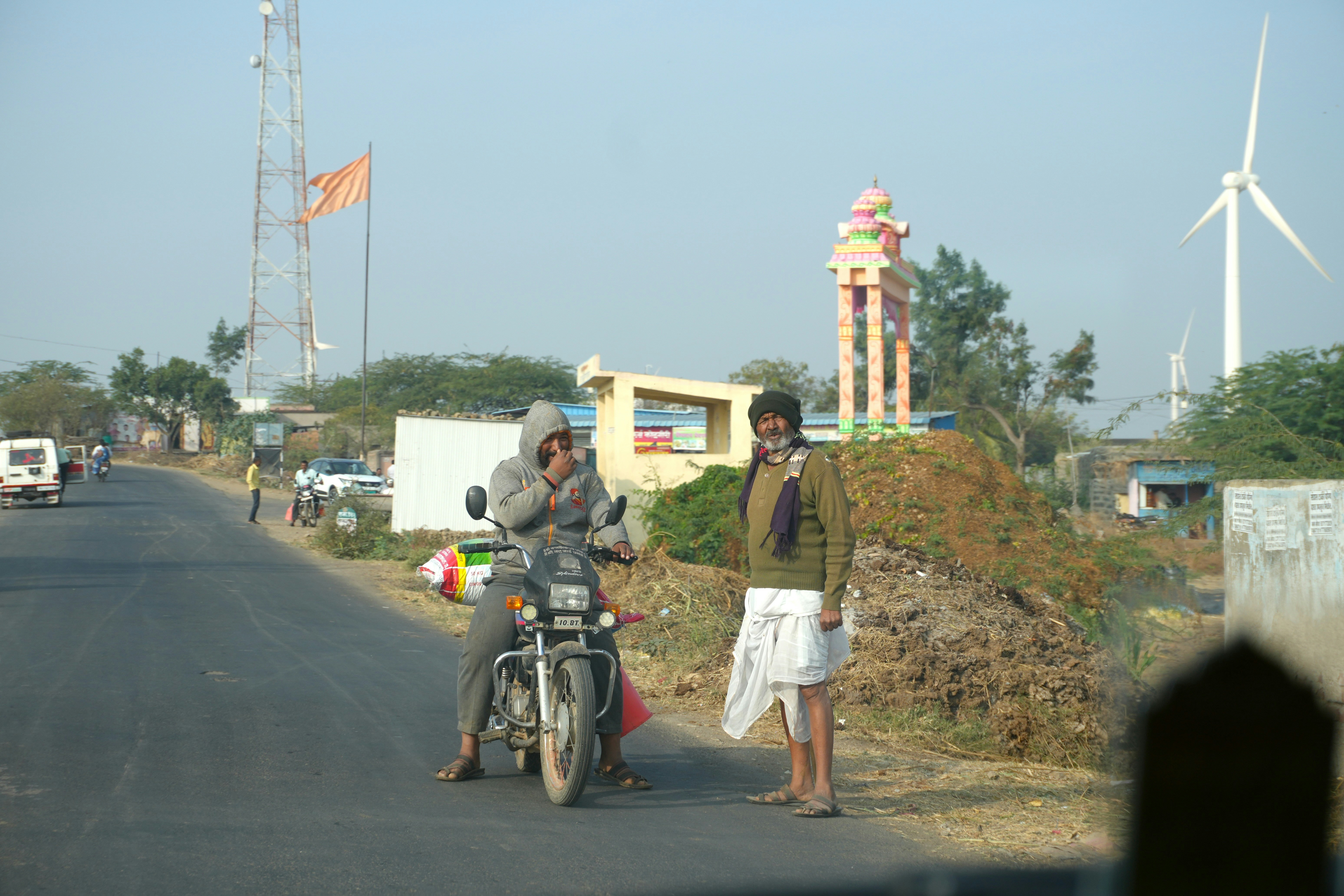 Two men by a motorcycle on a rural road, one sitting on the bike and the other standing beside, with wind turbines and a small shrine visible in the background.