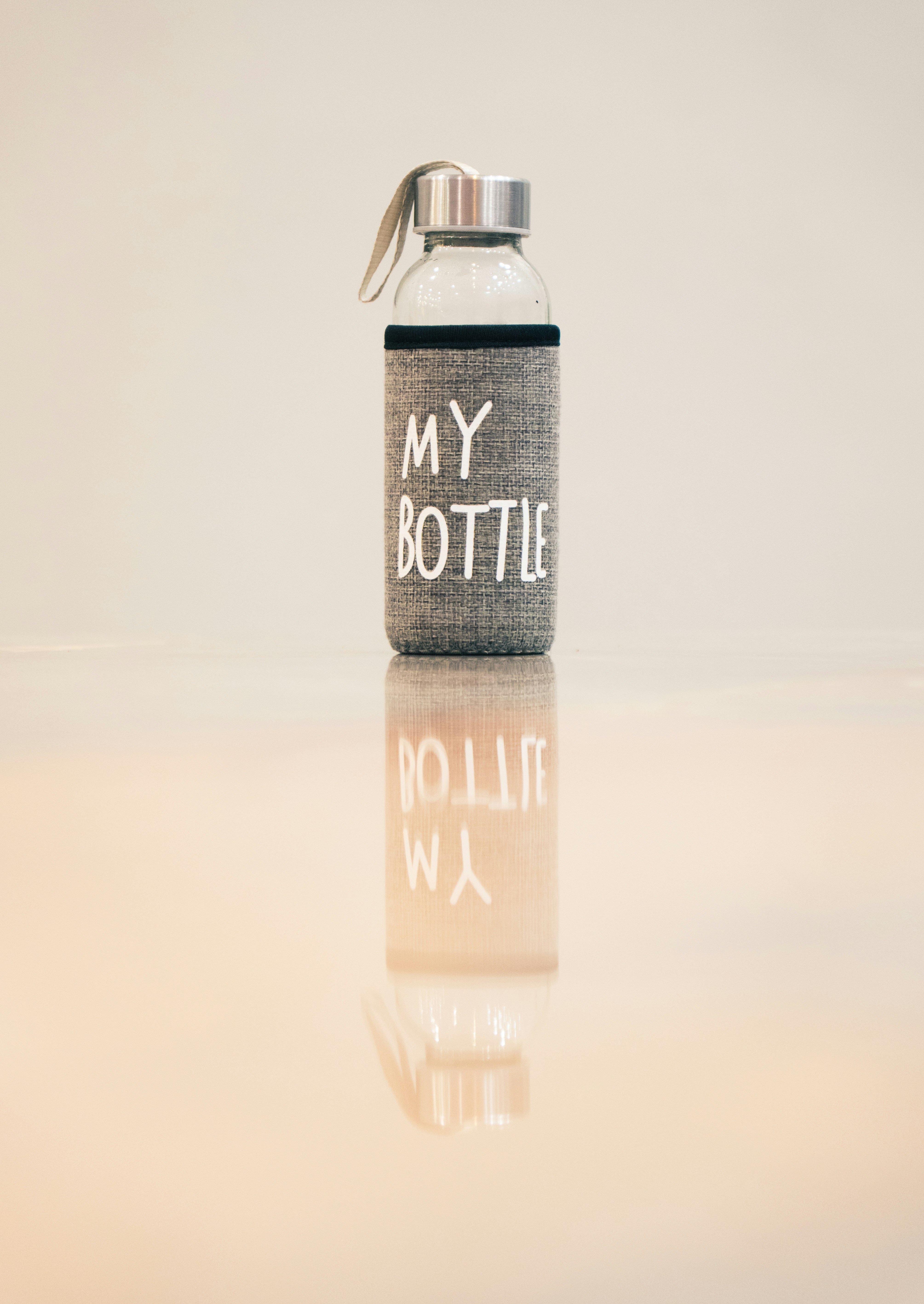 A glass water bottle wrapped in fabric sits on a reflective surface, with soft, neutral tones and a calm, minimal mood.