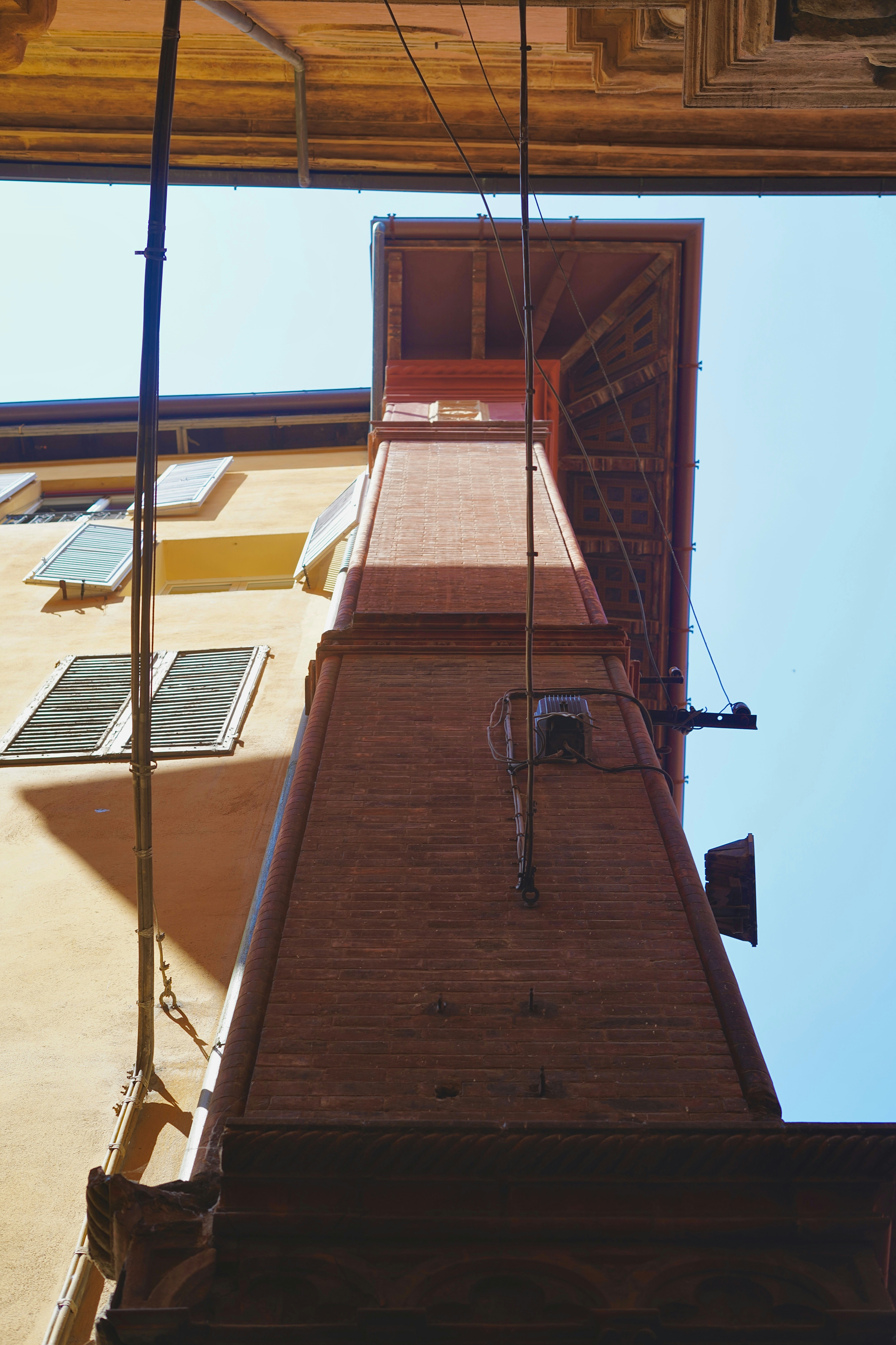 Brick tower rising against a clear blue sky with scaffolding lines and architectural details. A vertical composition emphasizing the tower’s height and the play of light and shadow.