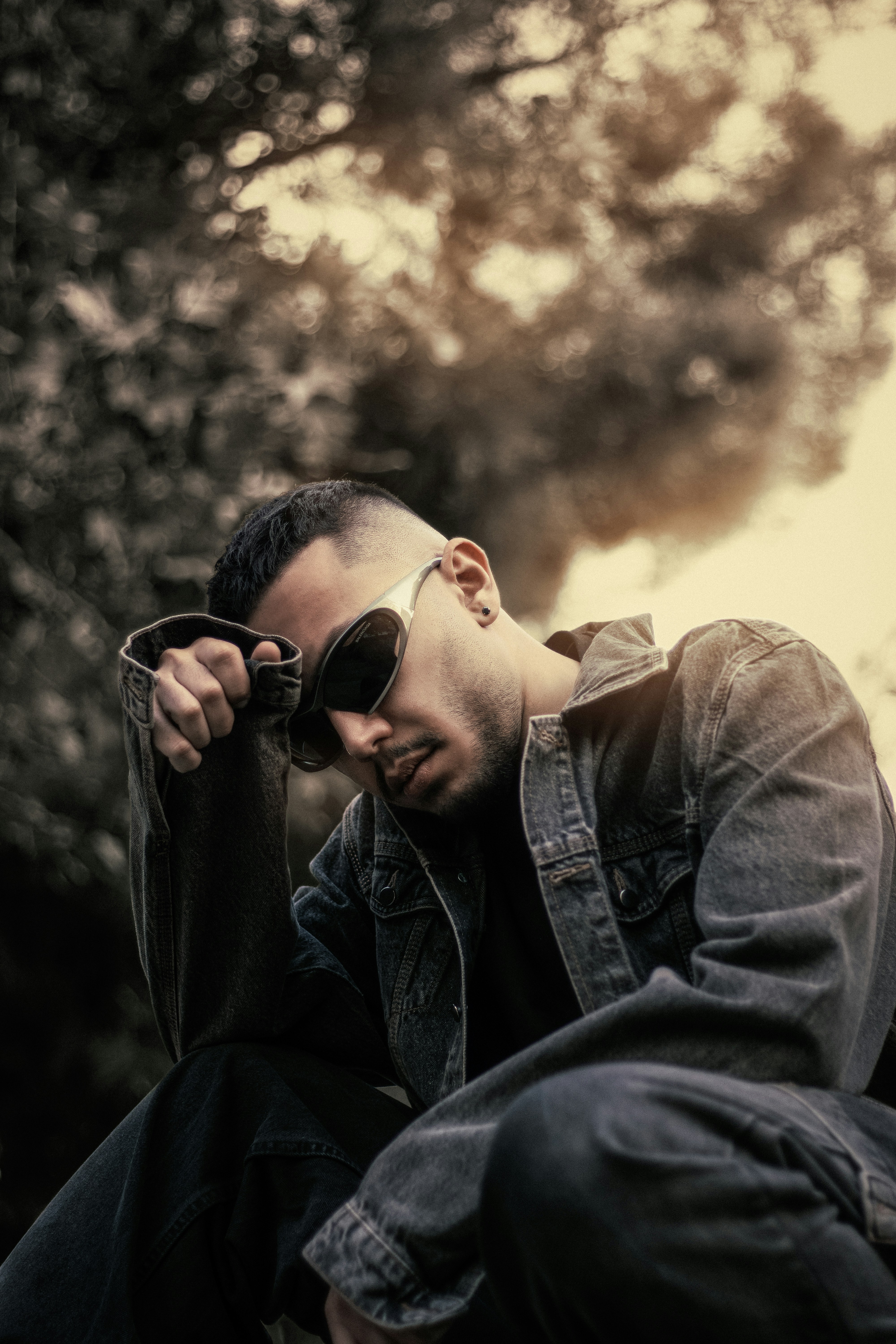Portrait of a man wearing sunglasses and a denim jacket, seated outdoors with a blurred leafy background, emphasizing mood and posture. Photograph appears as a moody, slightly retro capture.