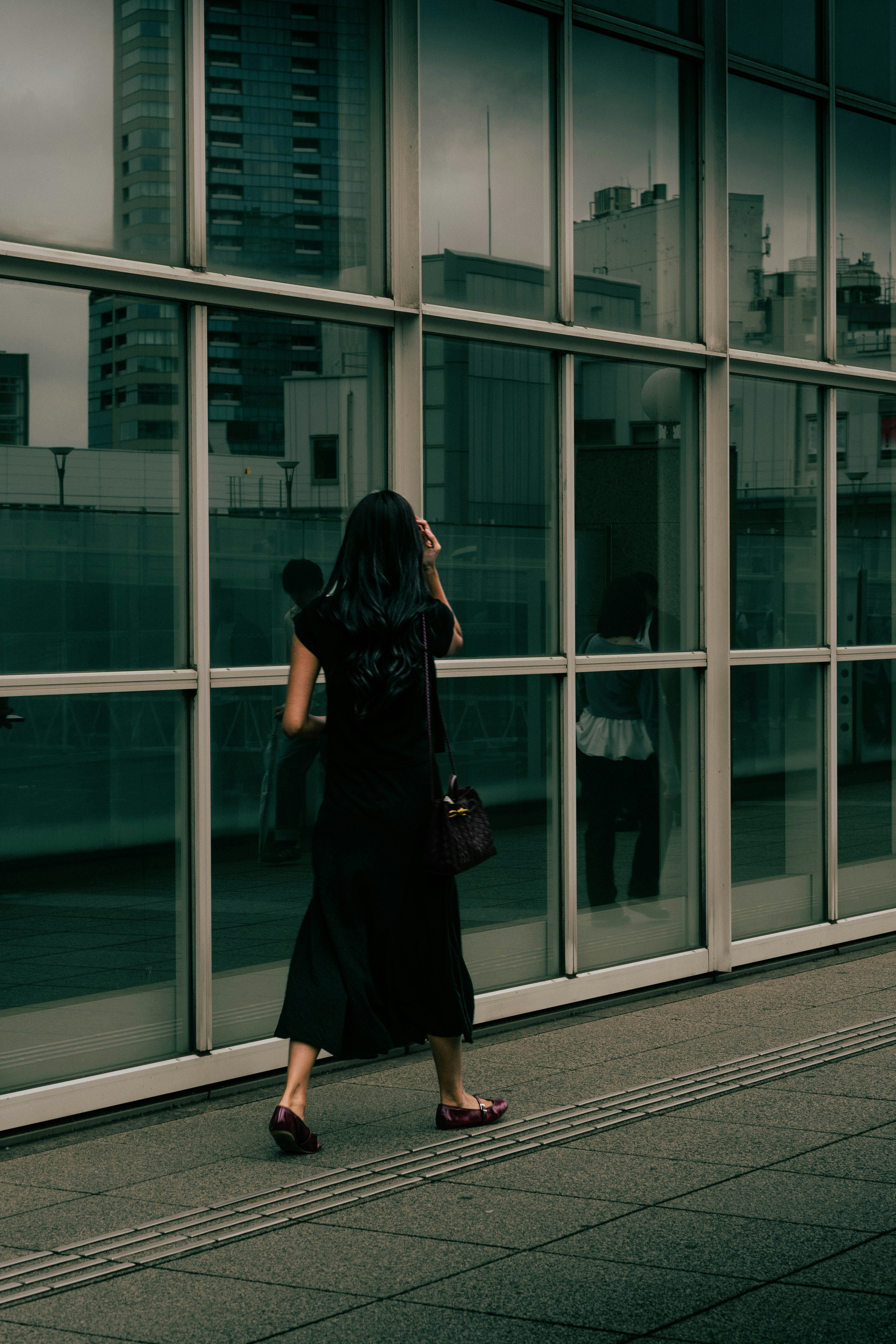 A woman in a black dress walks along a glass-fronted urban building, her silhouette reflected in the windows. The scene emphasizes modern architecture and street-level activity.