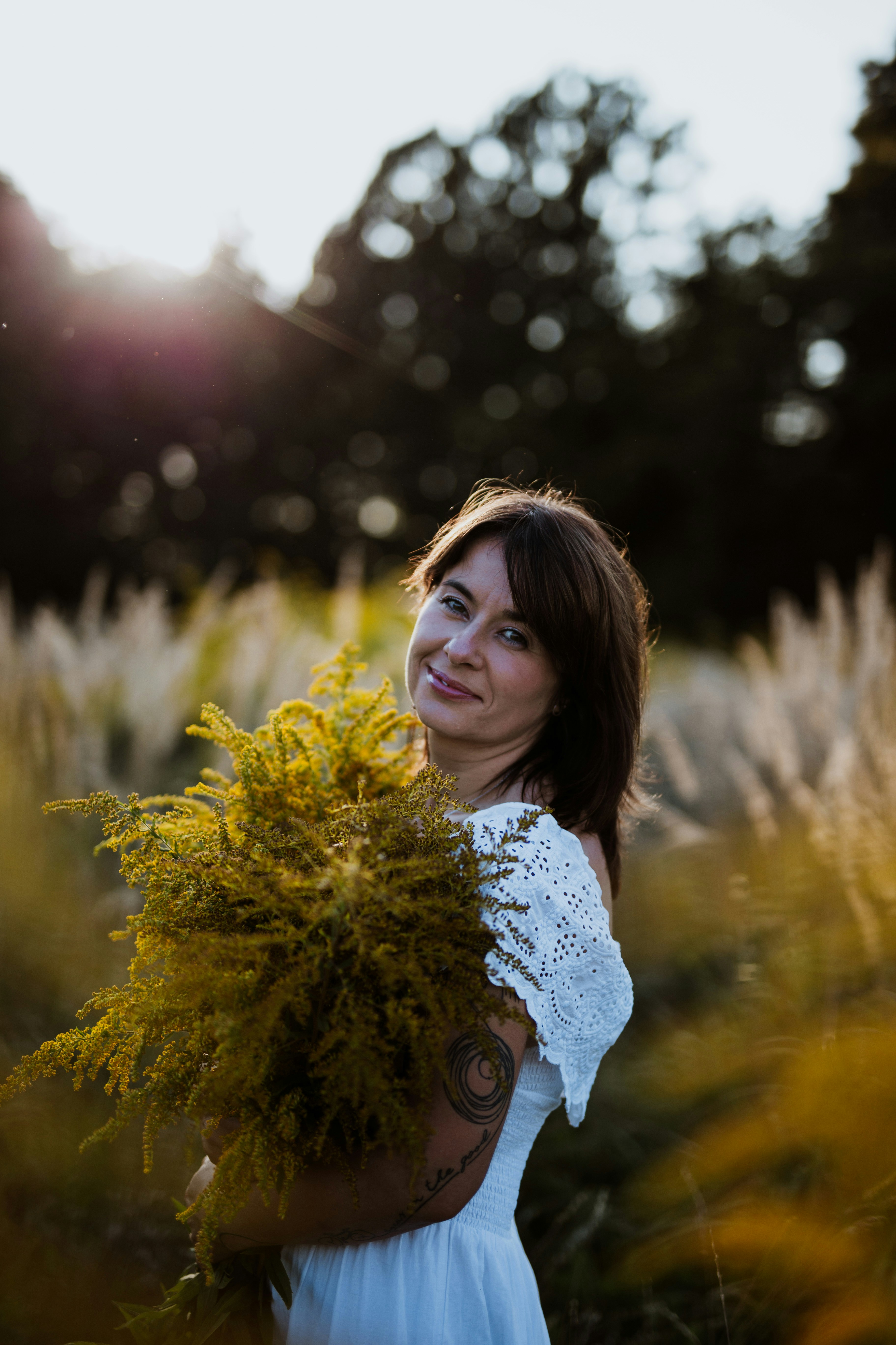 Portrait of a woman in a light blue dress holding a yellow bouquet, standing in a sunlit field with soft bokeh and trees in the background.
