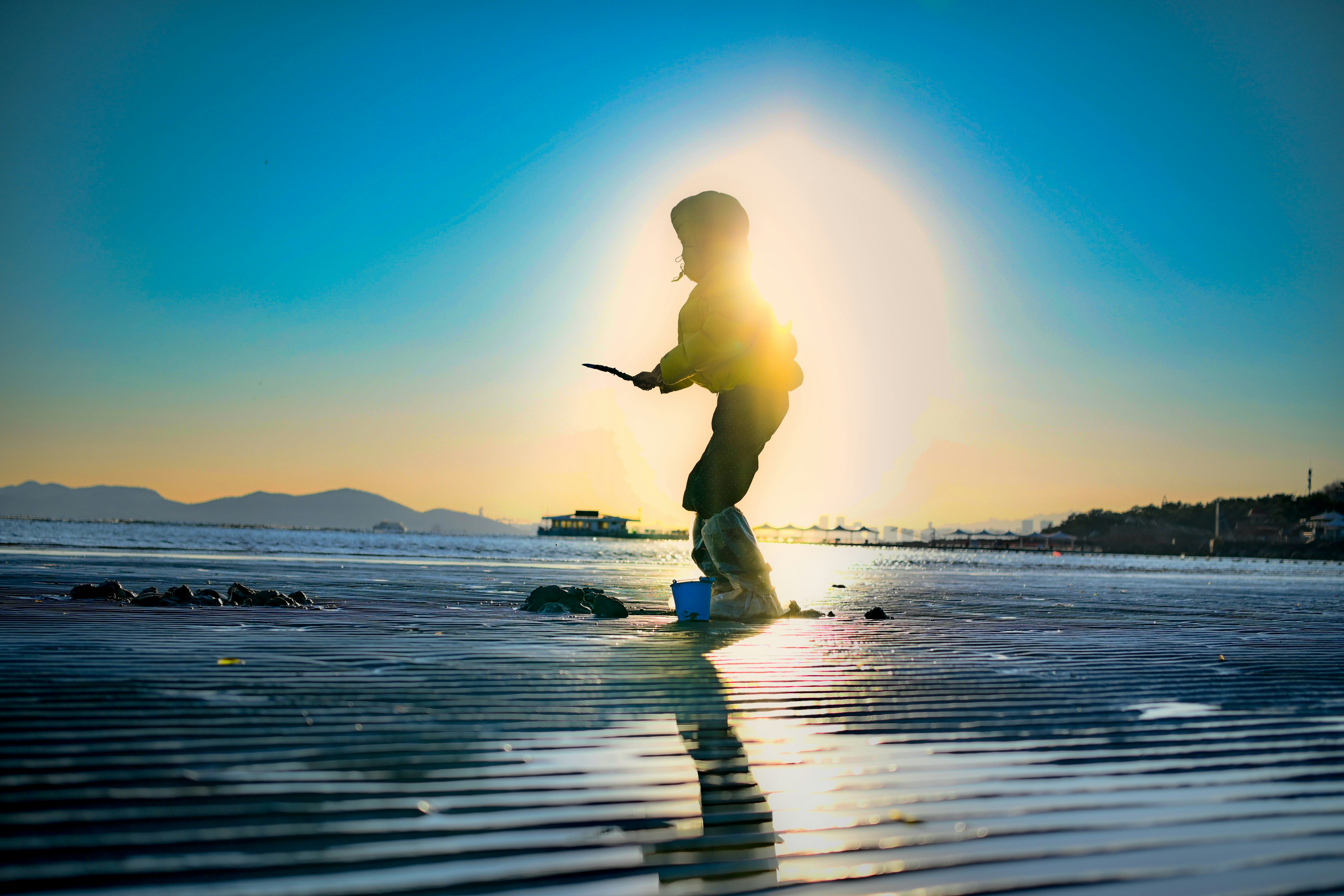 A child stands on wet beach boards at sunset, holding an object, with the sun halo behind creating a dramatic silhouette and reflective water patterns.