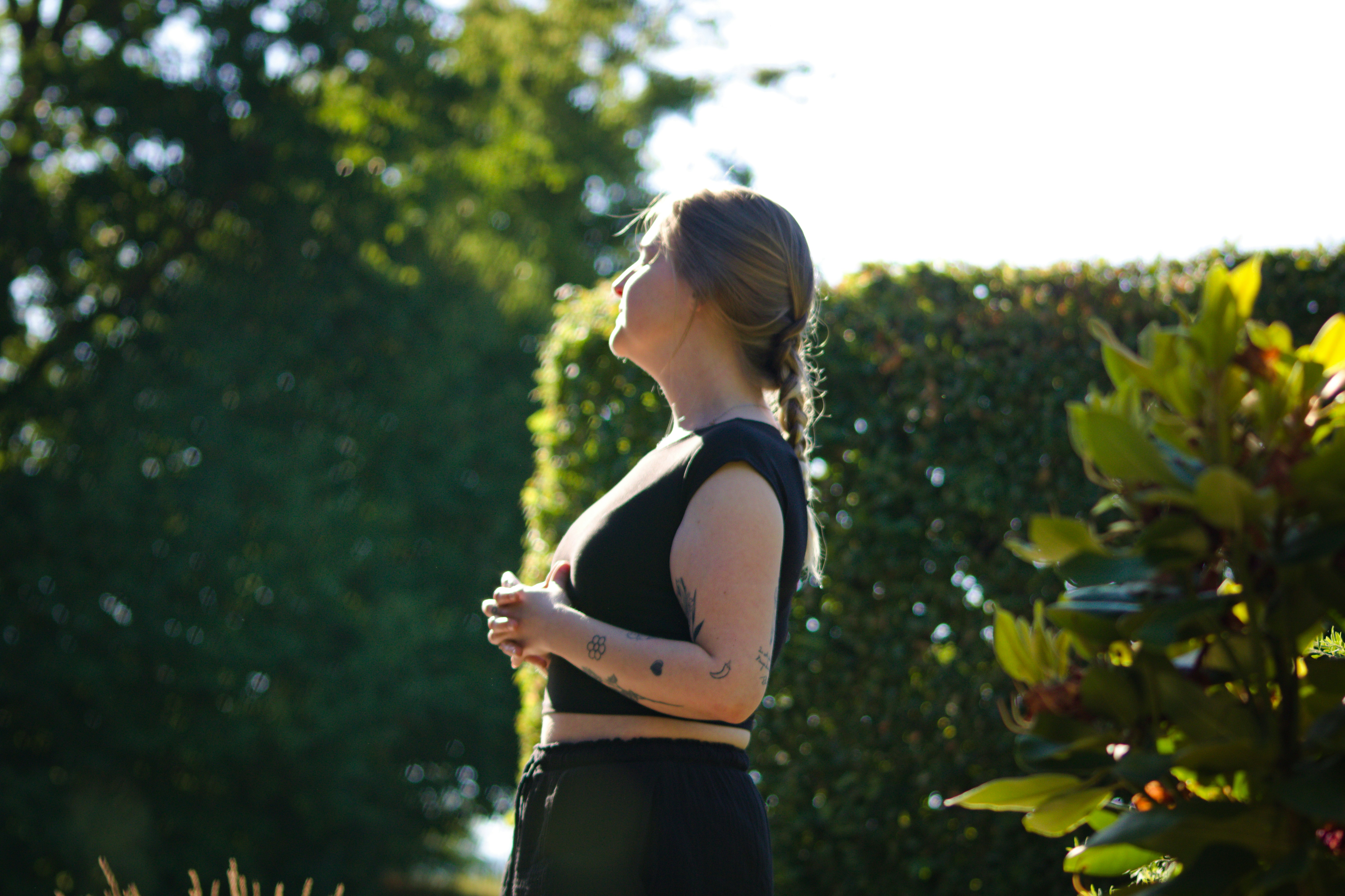 Woman stands sideways in a sunlit garden, head tilted toward the light, with greenery and a hedge in the background.