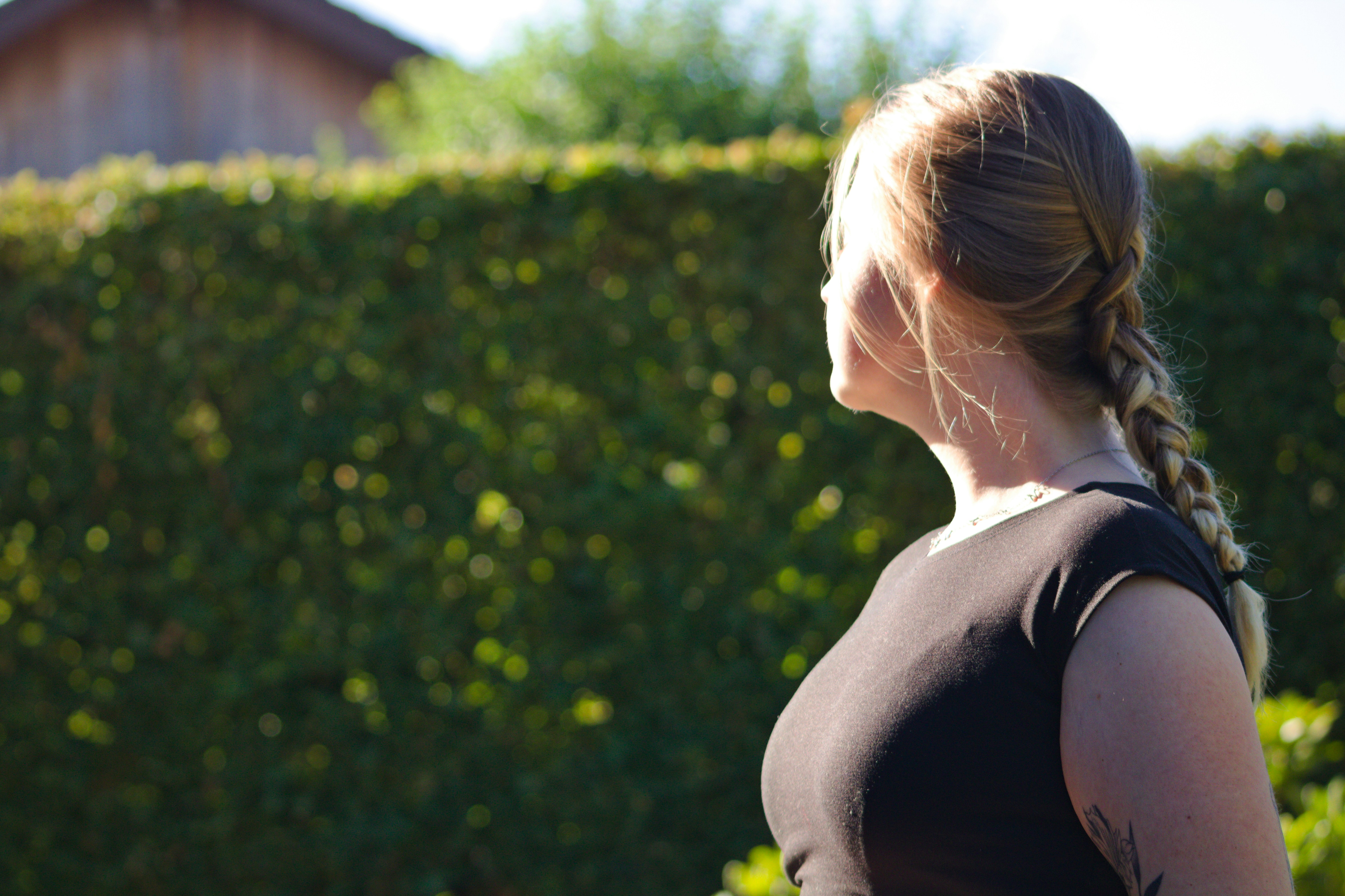 Sunlit profile of a woman with a braided ponytail wearing a black top, standing against a sunlit hedge; the scene emphasizes natural light and a shallow background blur.