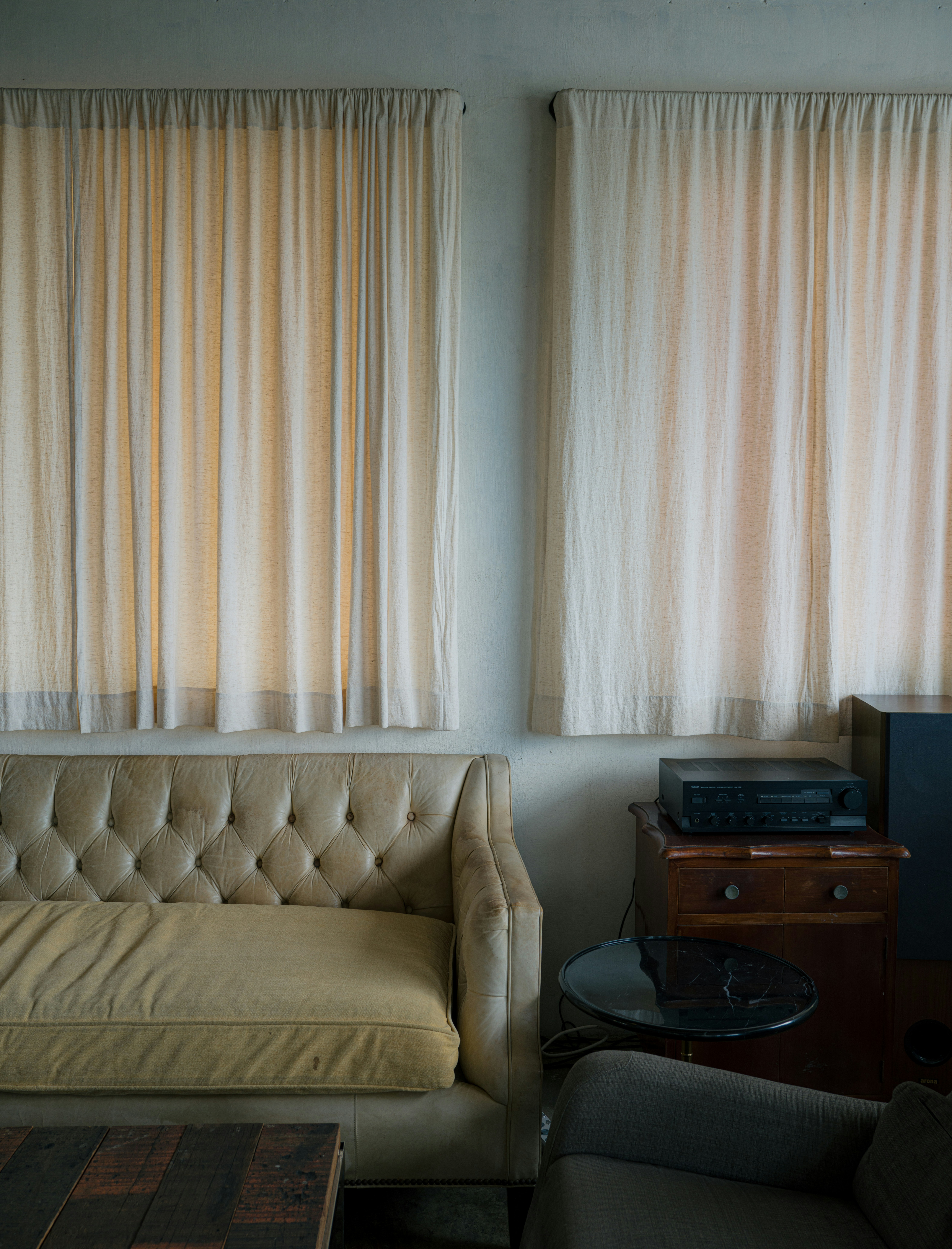 Muted living room scene with a tufted sofa, wood side table, and vintage audio components beneath cream curtains. A quiet, intimate interior study of light, texture, and midcentury design.