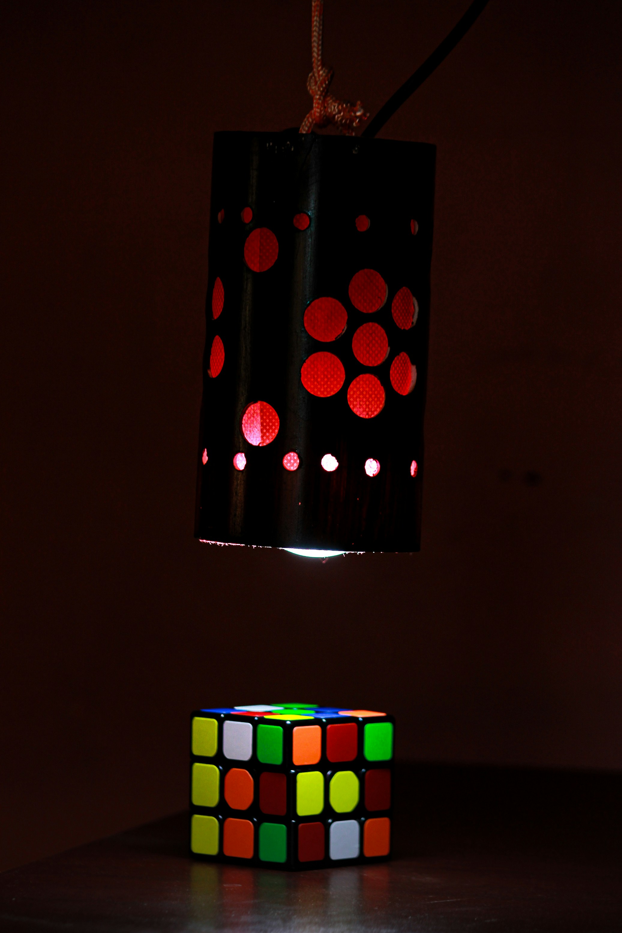 Dimly lit scene featuring a cylindrical lantern with red circular cutouts suspended above a Rubik’s Cube on a dark surface.