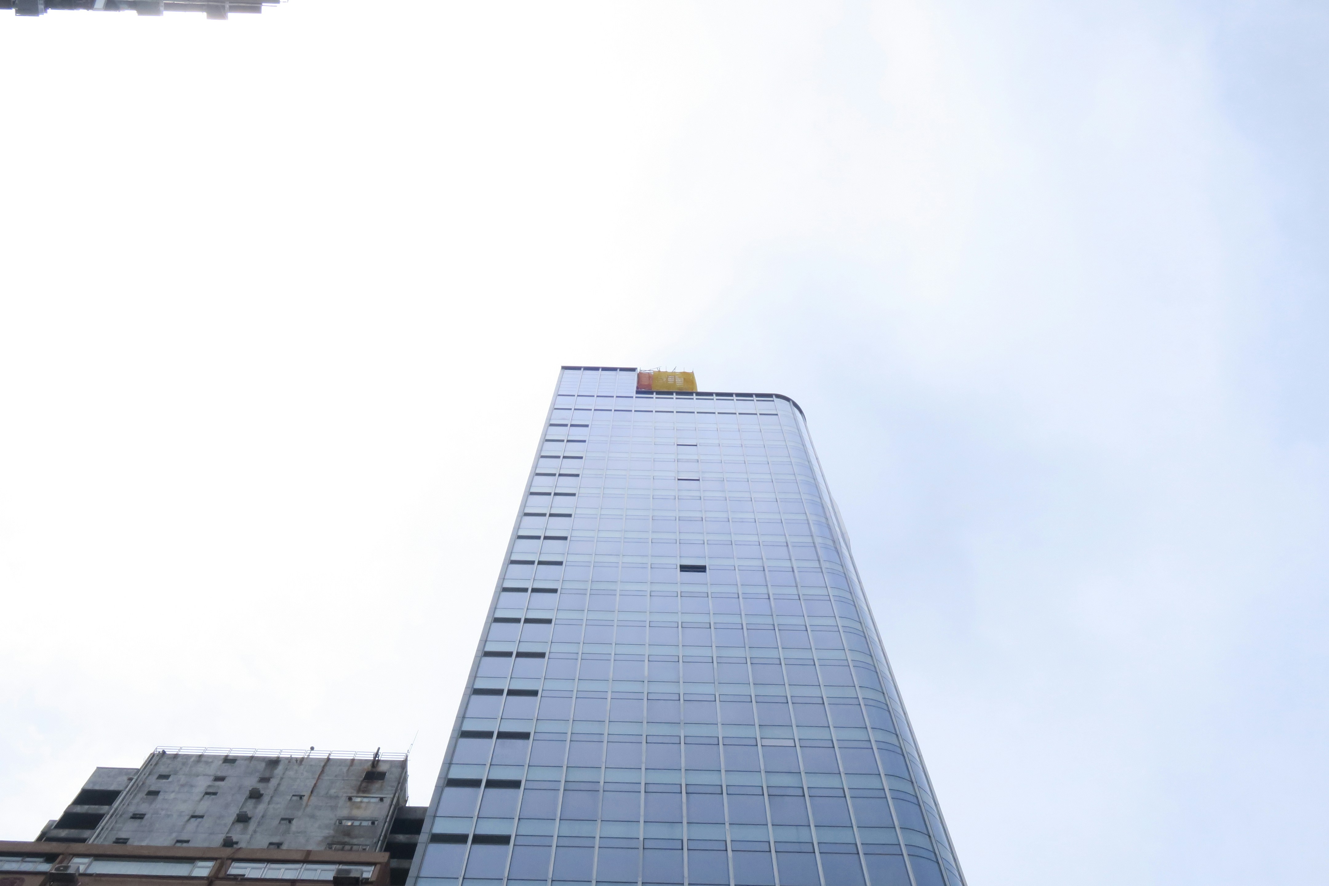 Tall glass skyscraper rising diagonally against a pale sky, with adjacent low-rise buildings at the base.