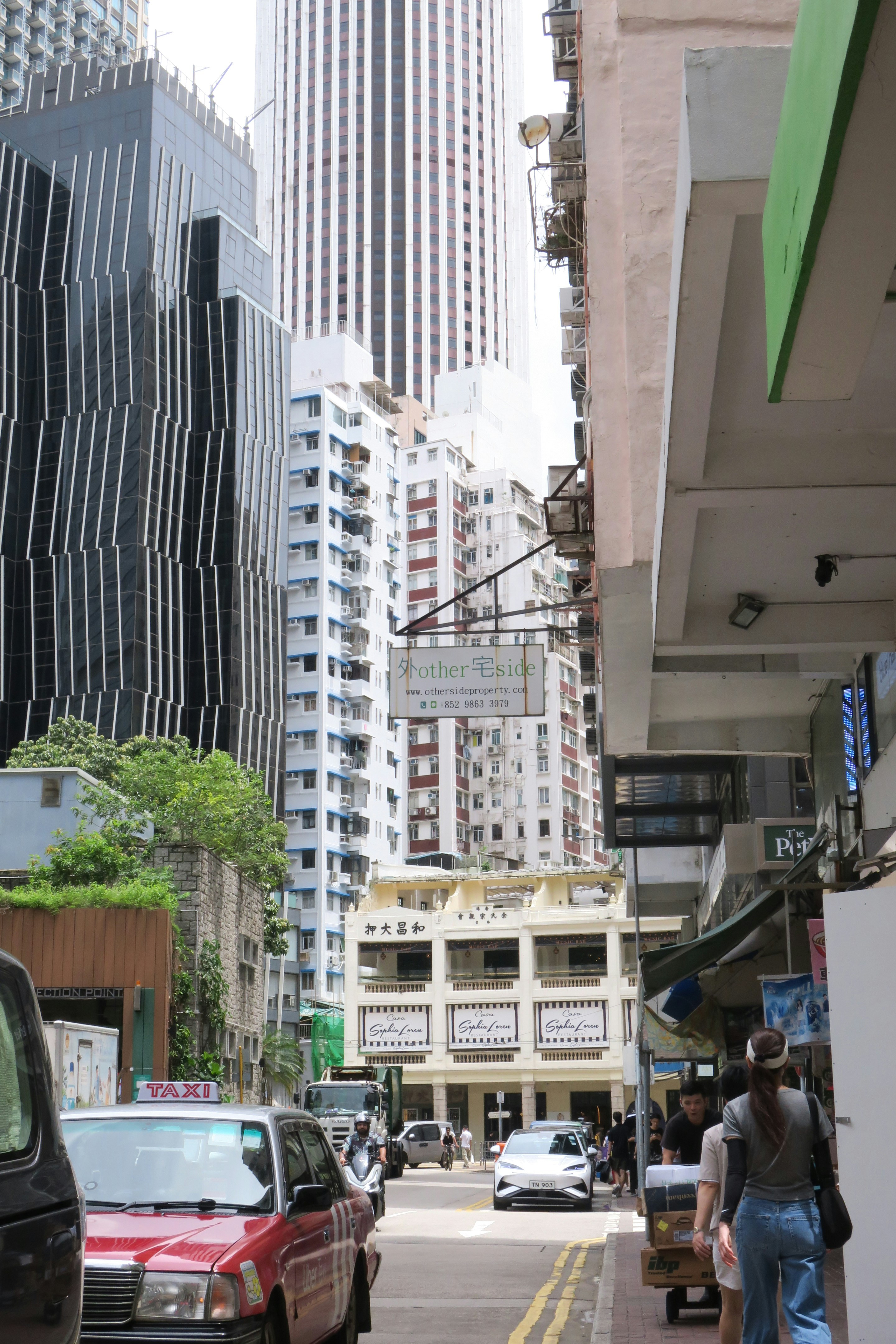 Bustling city street with parked cars, pedestrians, and high-rise buildings lining the avenue; a mix of modern glass towers and older structures creates a dense urban canyon.