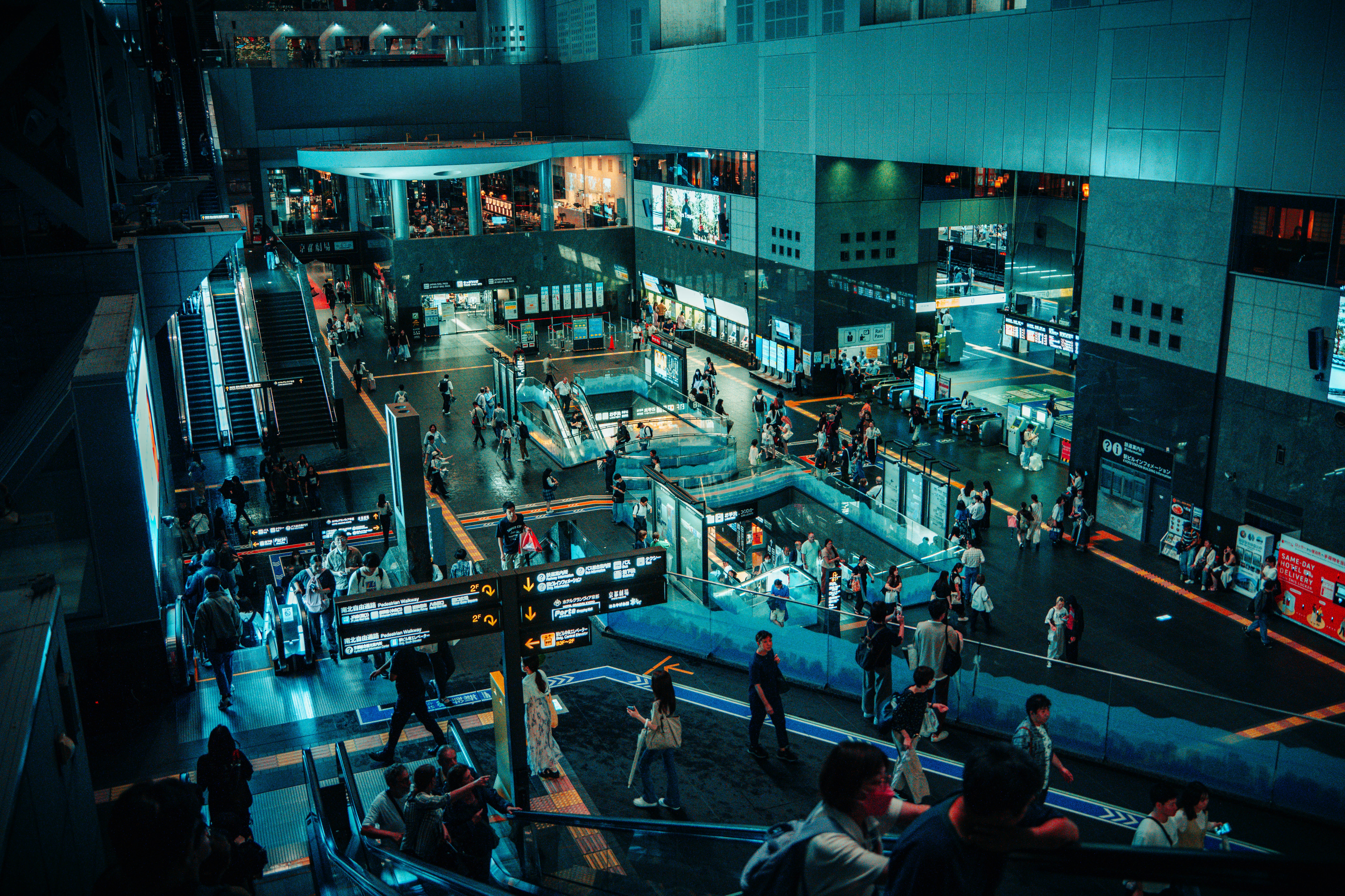 Busy multi-level atrium with people moving along walkways and escalators, illuminated by blue-green lighting and retail storefronts; a dynamic interior scene with varied depth and reflections.