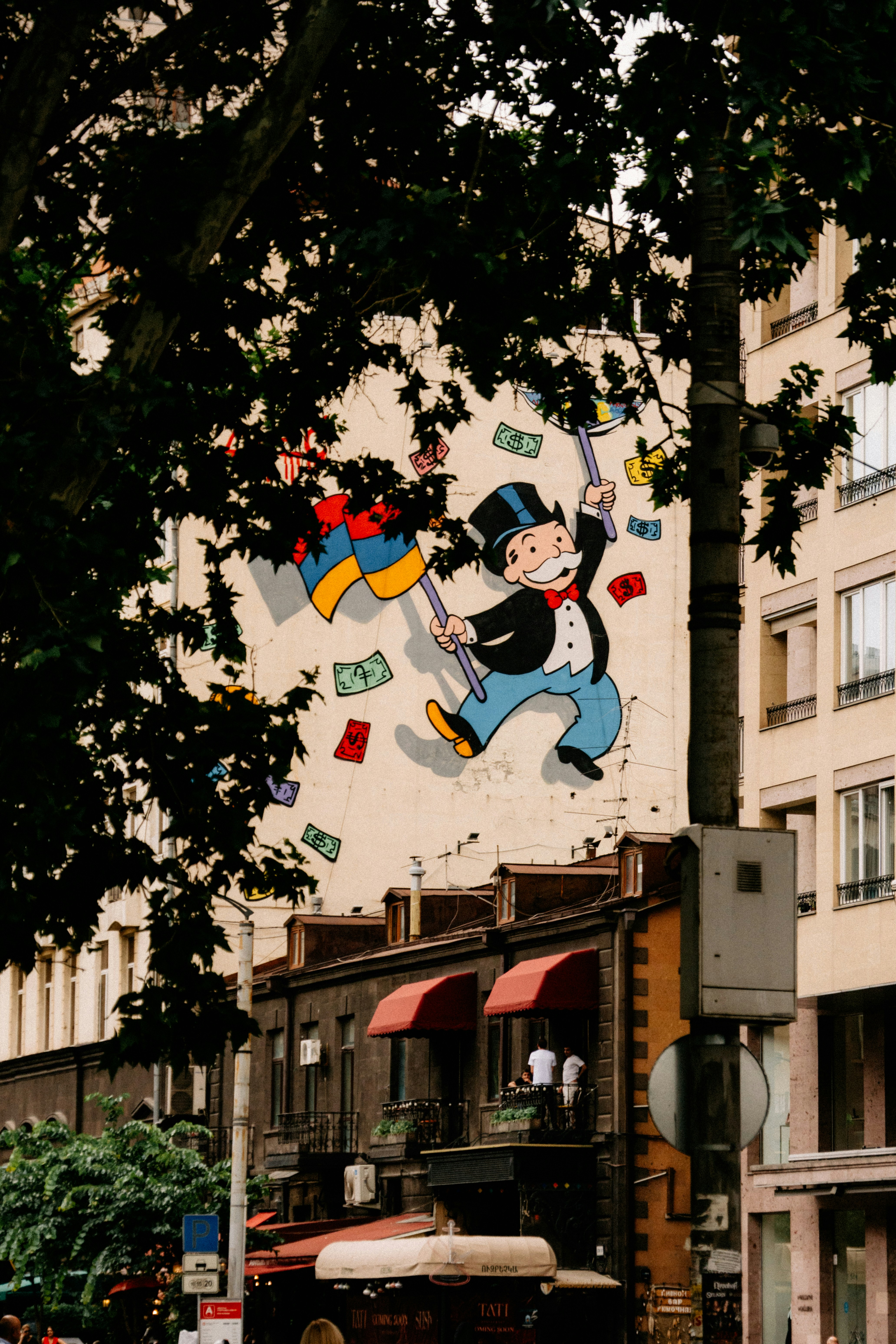 Colorful mural of a marching cartoon figure on a building wall amid city blocks; foreground includes storefronts and trees, suggesting a lively urban scene.
