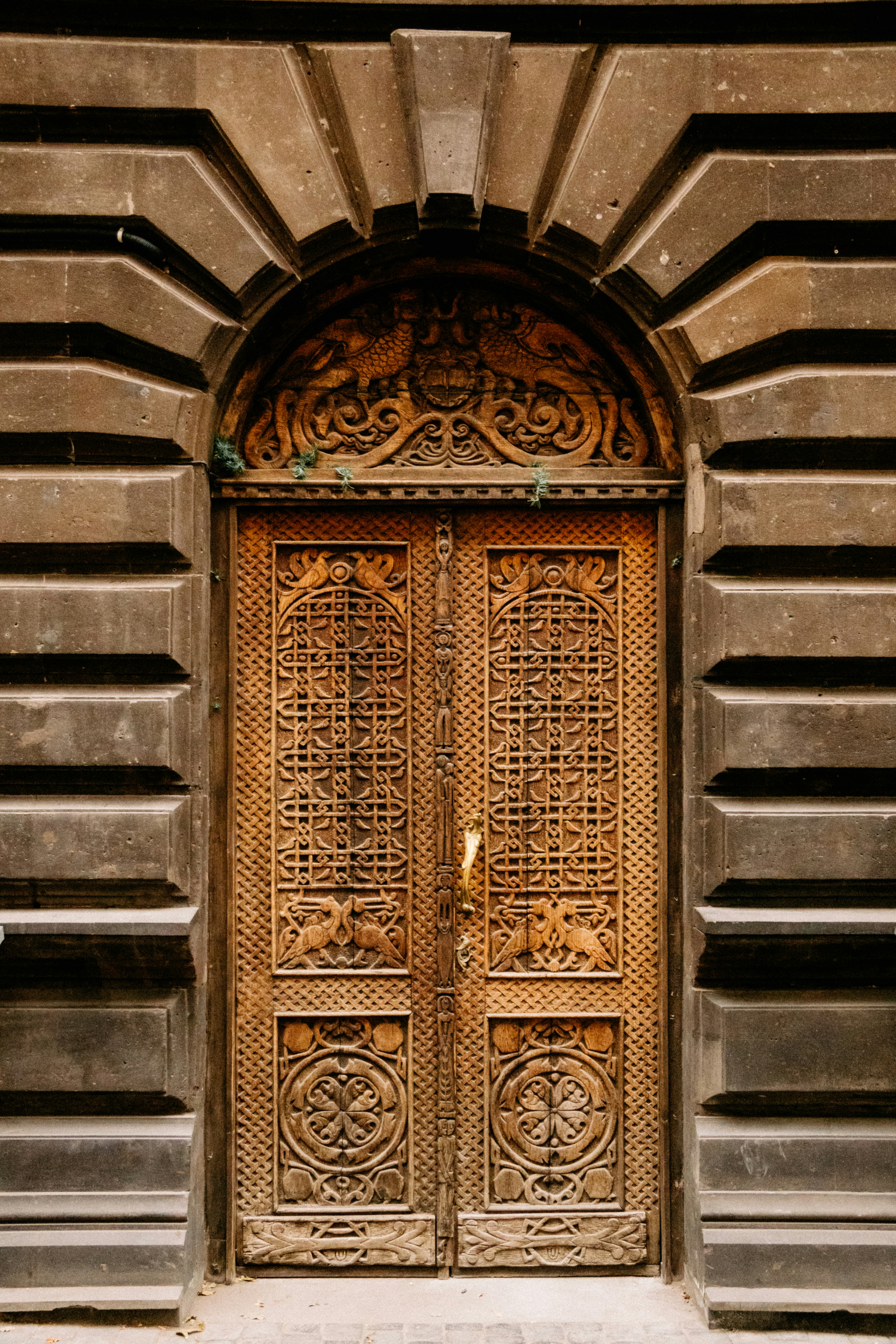 Intricately carved wooden double door set within a heavy stone facade, featuring arabesque lattice panels and ornamental reliefs under a rounded arch.