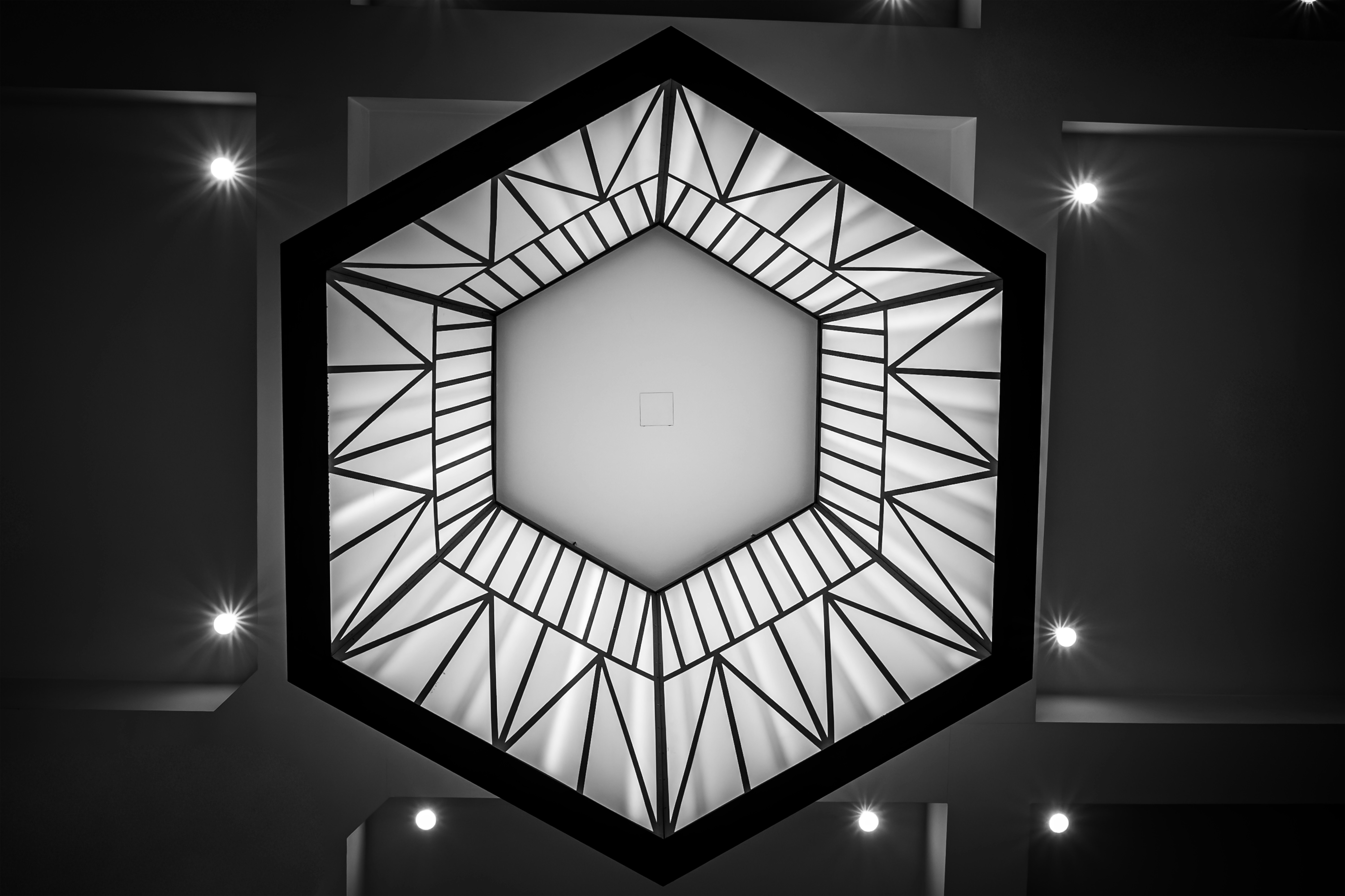 Monochrome interior ceiling with a hexagonal central light frame, geometric patterns radiating outward. Minimalist, architectural composition highlighting symmetry and perspective.