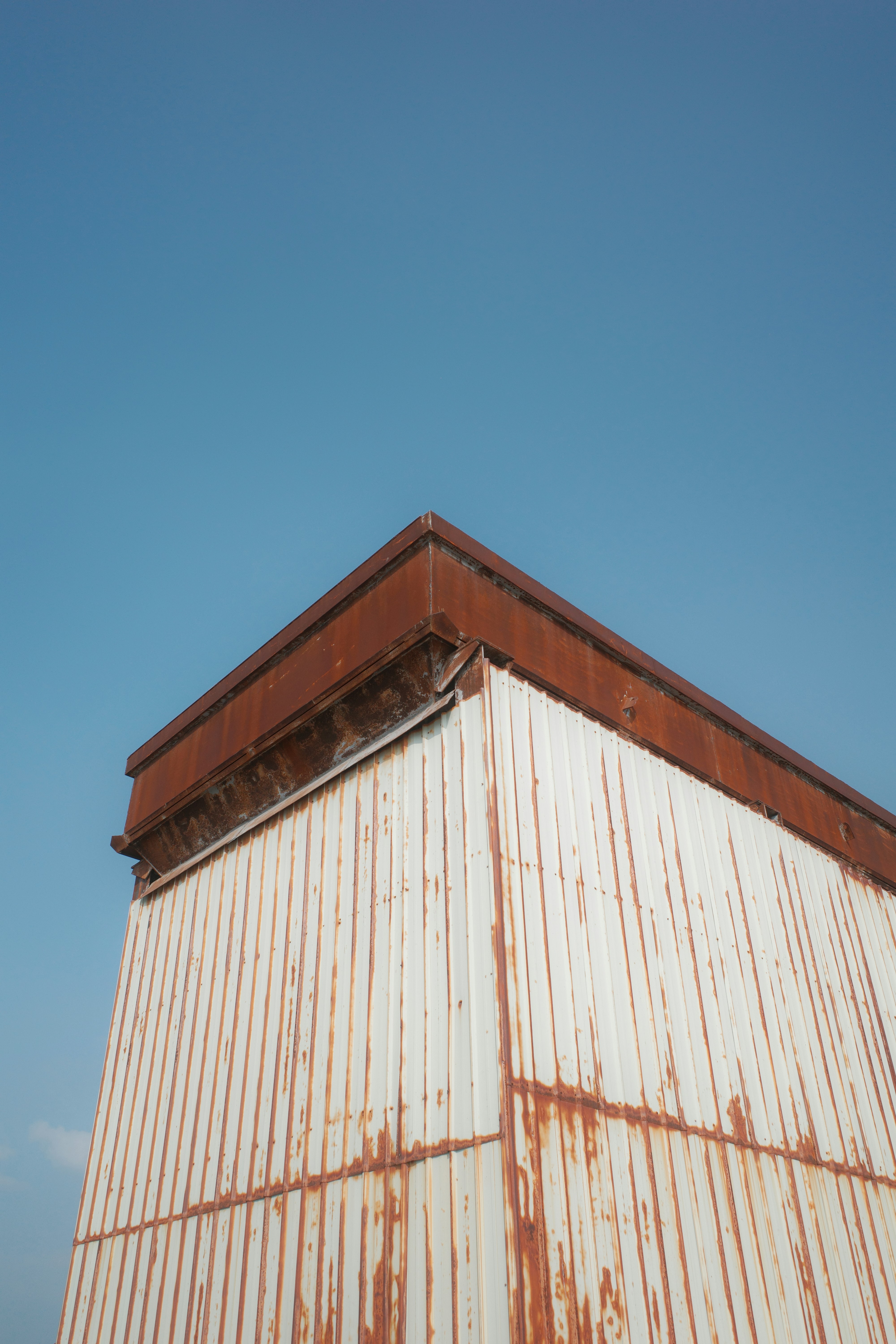Rusty metal shed corner against a clear blue sky, with vertical panels and weathered rust accents. A minimalist architectural detail highlighted for texture and color.