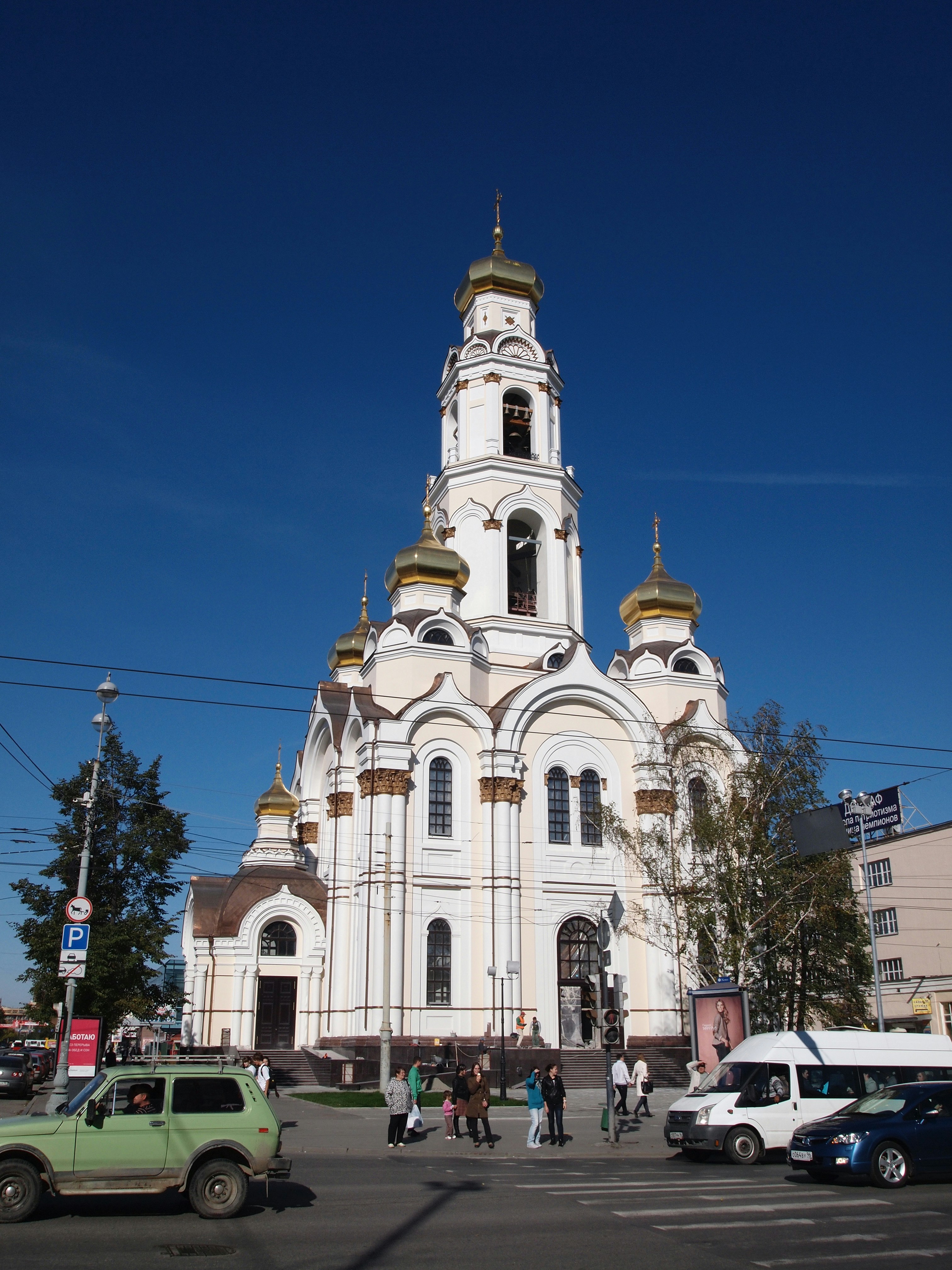 White Orthodox cathedral with golden domes rises above street level, pedestrians and vehicles at the entrance, clear blue sky backdrop.