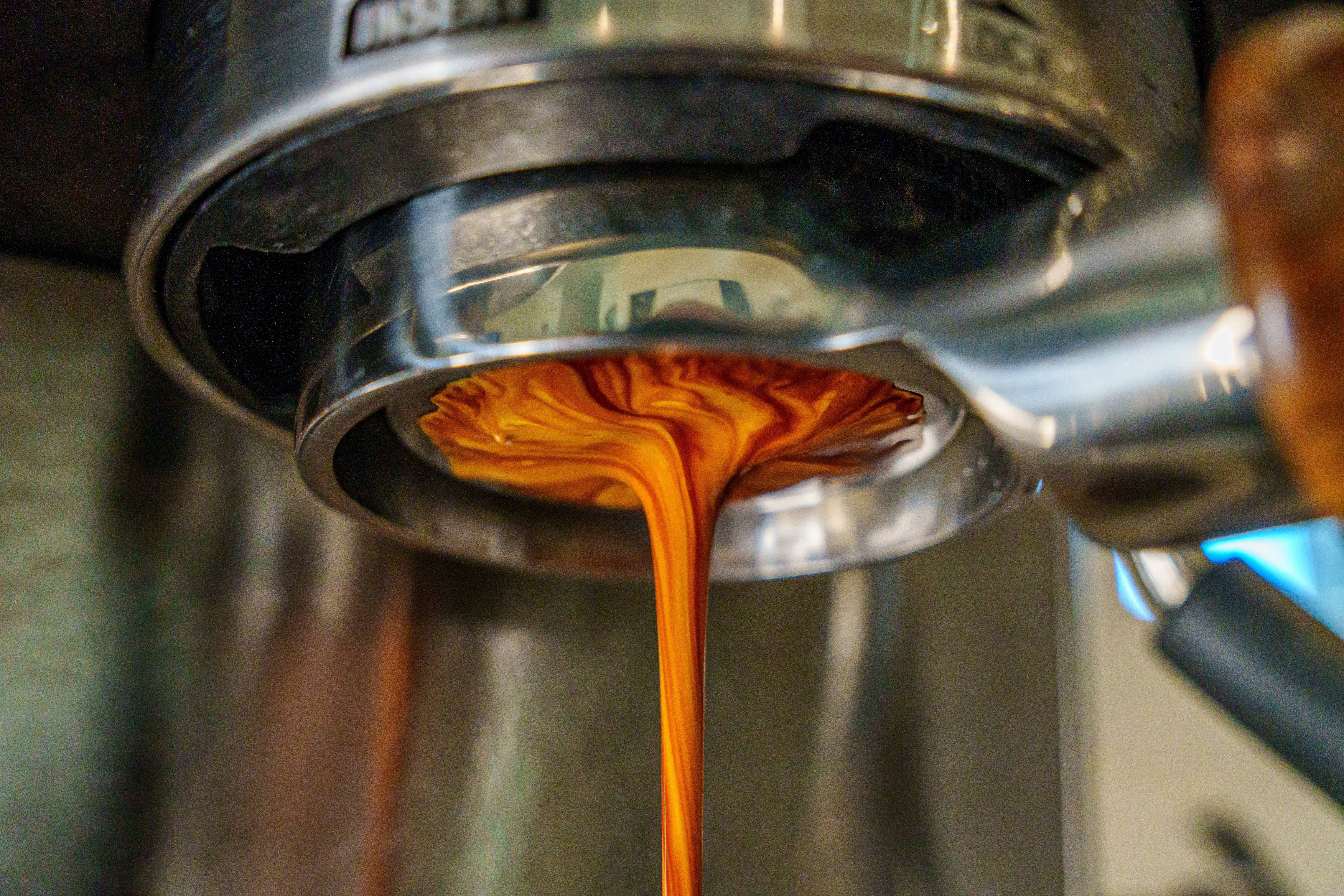 Close-up of hot espresso pouring from a portafilter into a cup, with metallic reflections and a hint of machine components in the background.
