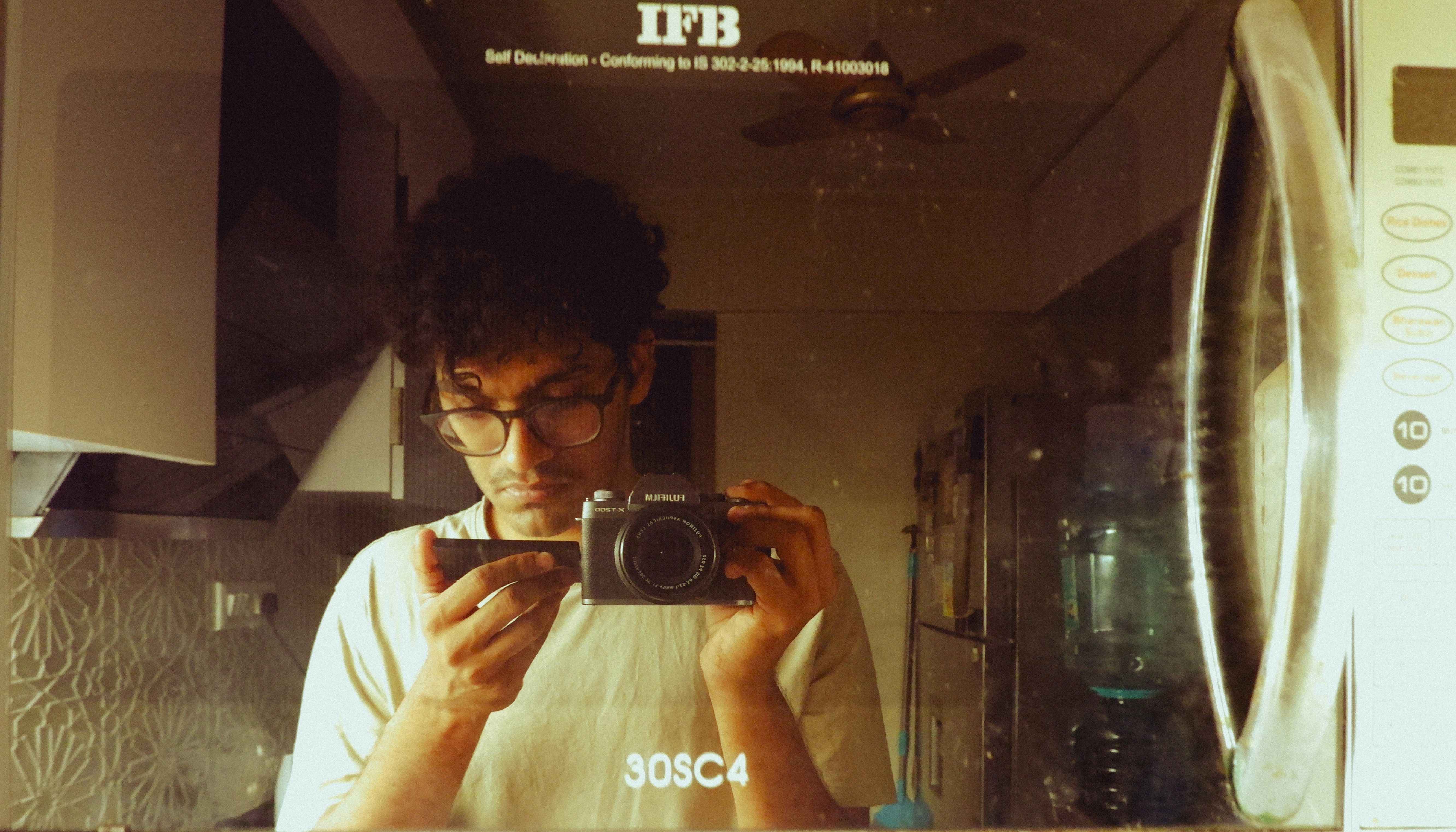 A young man with glasses holds a camera, capturing a self-portrait in a reflective surface; the scene includes a visible phone and camera, suggesting a casual, indoor setting. This is a photo taken through a mirror, likely a storefront or appliance area.